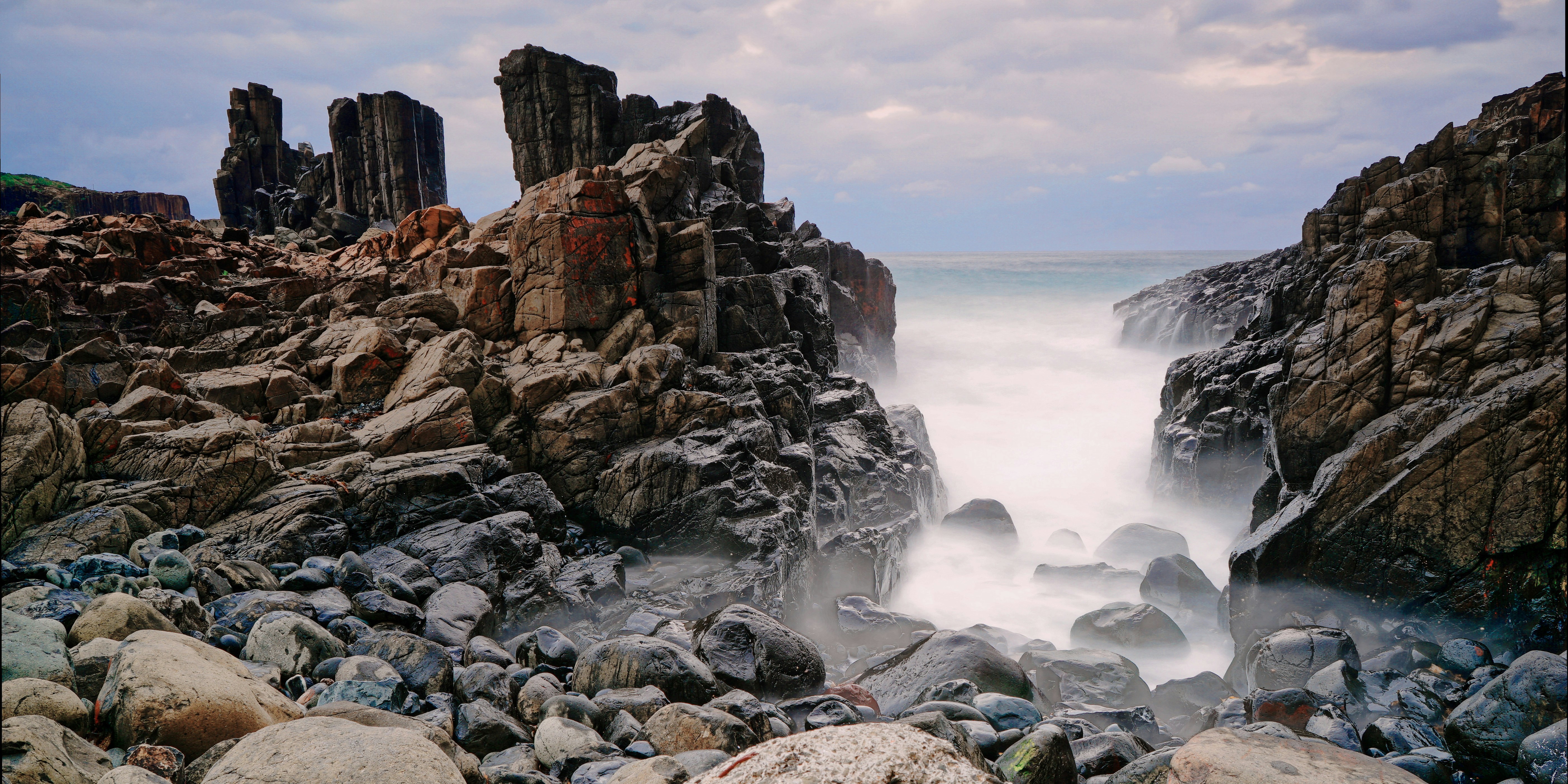 Waves crash against rocky cliffs under a cloudy sky, creating a misty seascape.