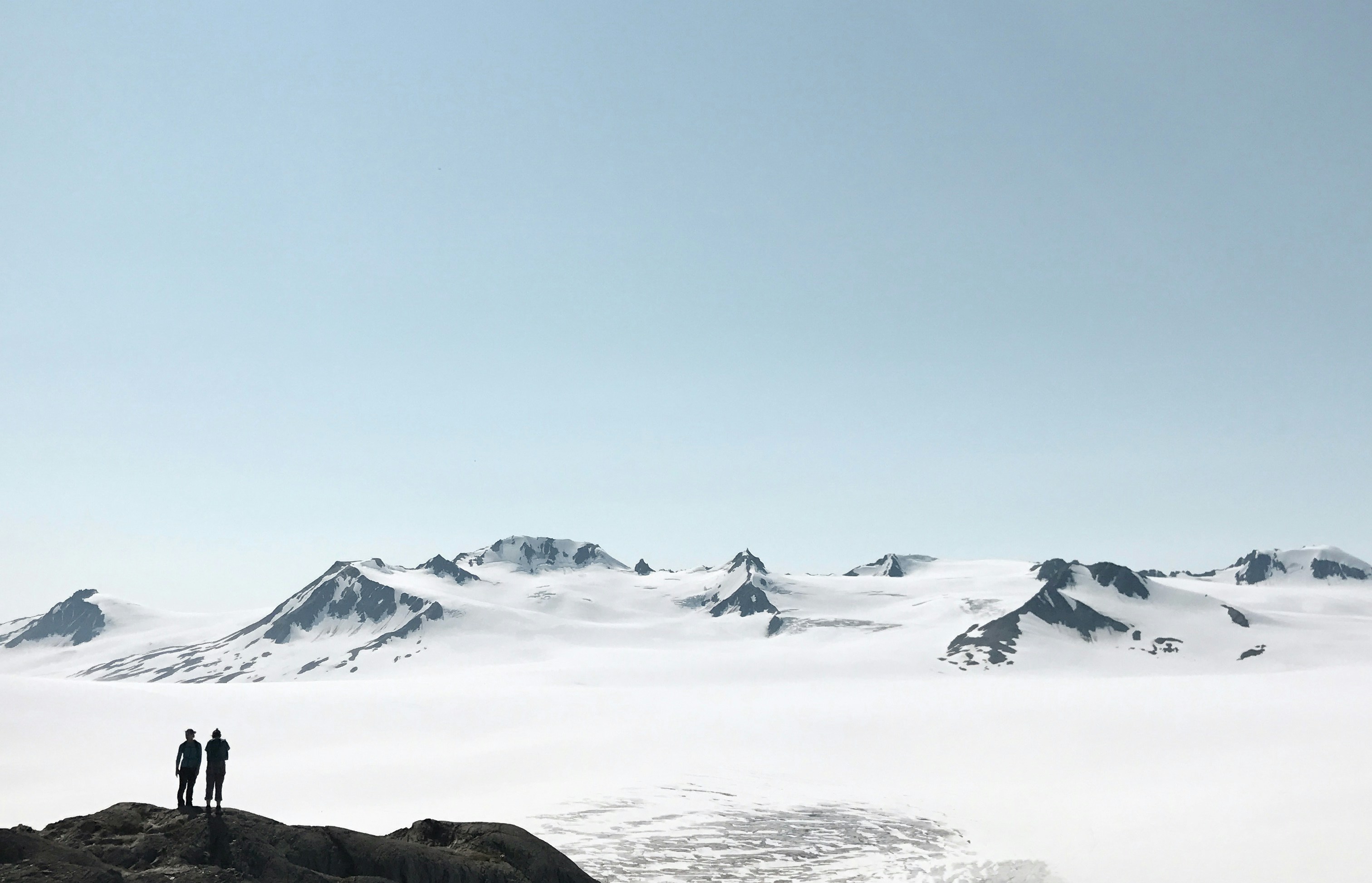 Two people stand on a rocky outcrop overlooking a vast snowy mountain range under a clear sky.