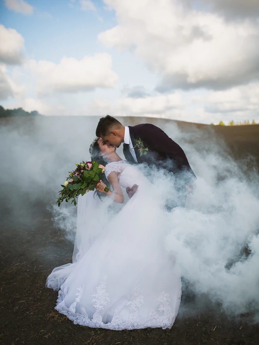 Bride and groom sharing a kiss outdoors, surrounded by drifting smoke under a cloudy sky. The bride holds a bouquet as the scene feels cinematic and romantic.