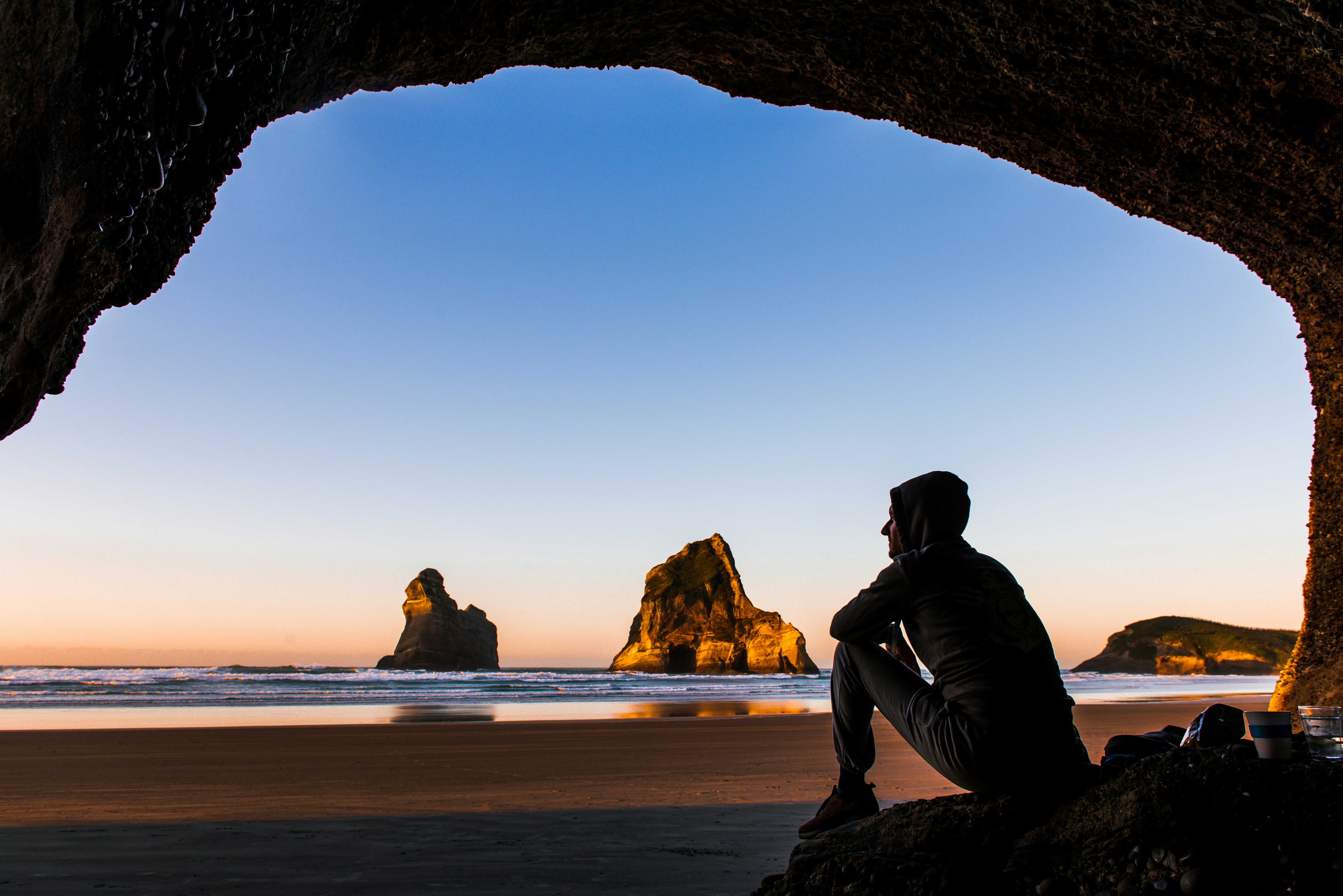 Silhouette of a person sitting in a cave, gazing at the ocean and rock formations during twilight.
