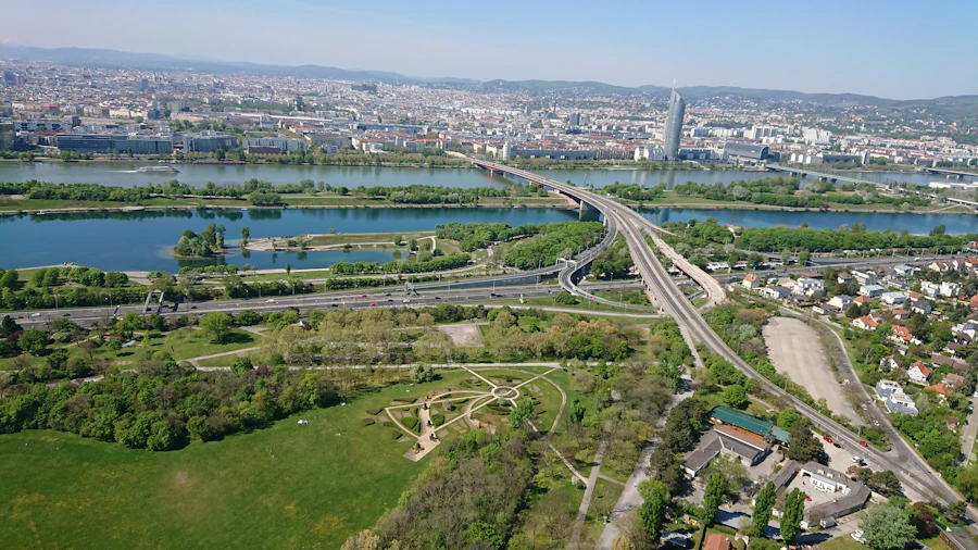 Aerial view of a city split by a wide river, with a sweeping bridge leading toward a dense skyline. Green parkland and roads fill the foreground under clear blue sky.