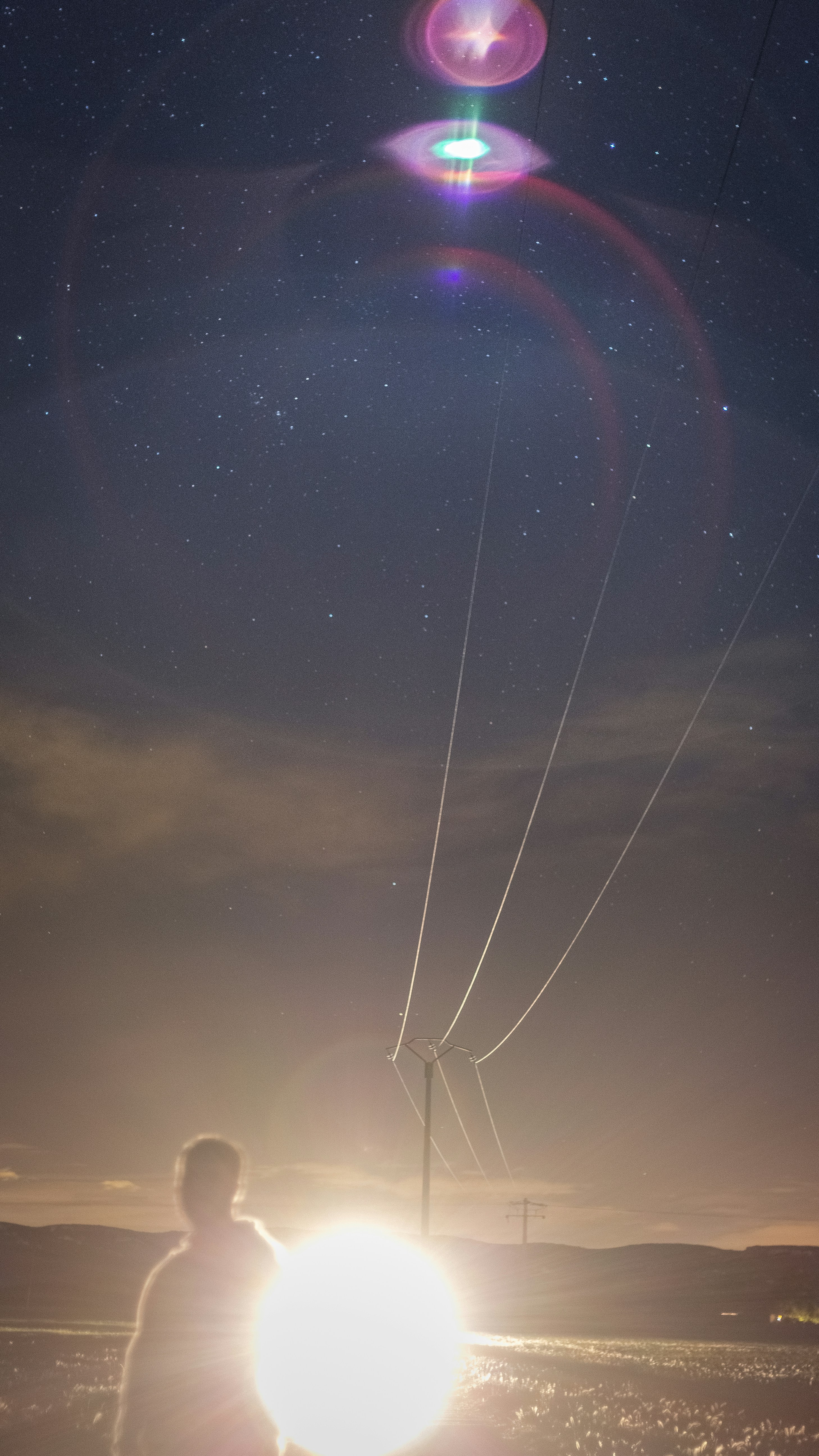 Silhouette of a person standing in a field, illuminated by a bright light source, with a starry night sky and power lines in the background.