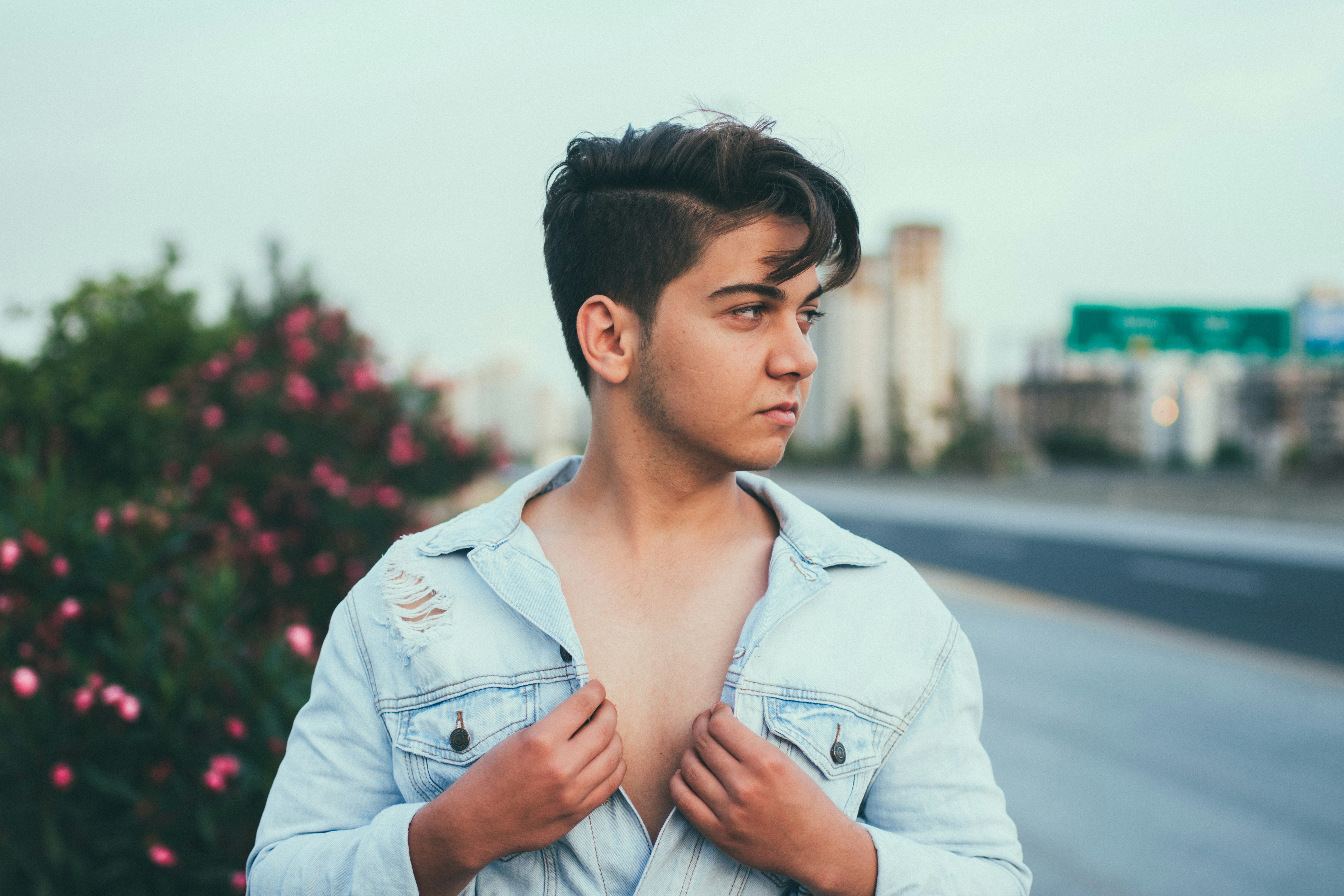 Young man in a denim jacket stands confidently against a blurred city backdrop, embodying a sense of self-expression and urban lifestyle.