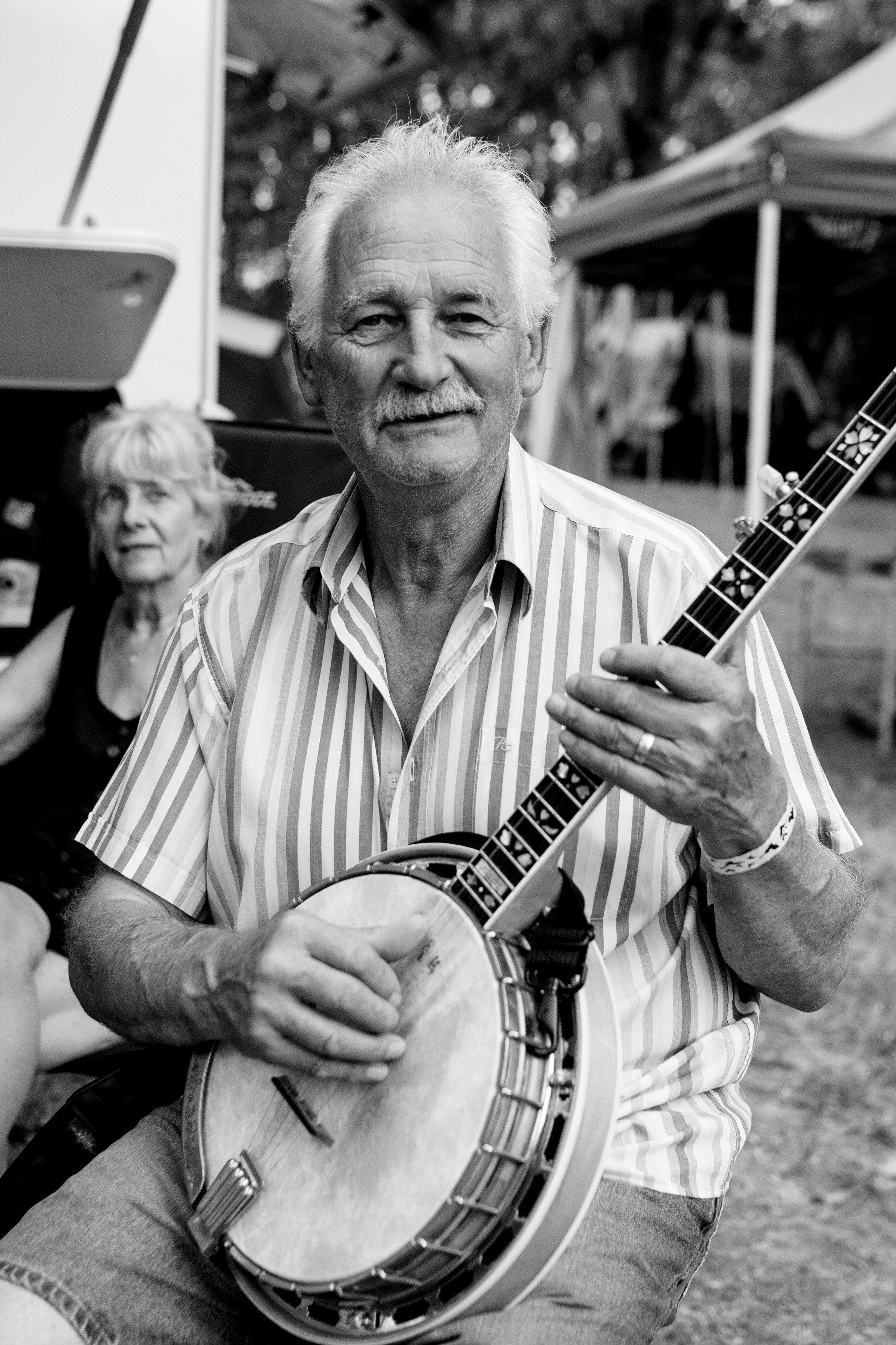An elderly man playing a banjo, showcasing his passion for music, while a woman sits in the background. The scene captures a moment of joy and connection at a lively gathering.