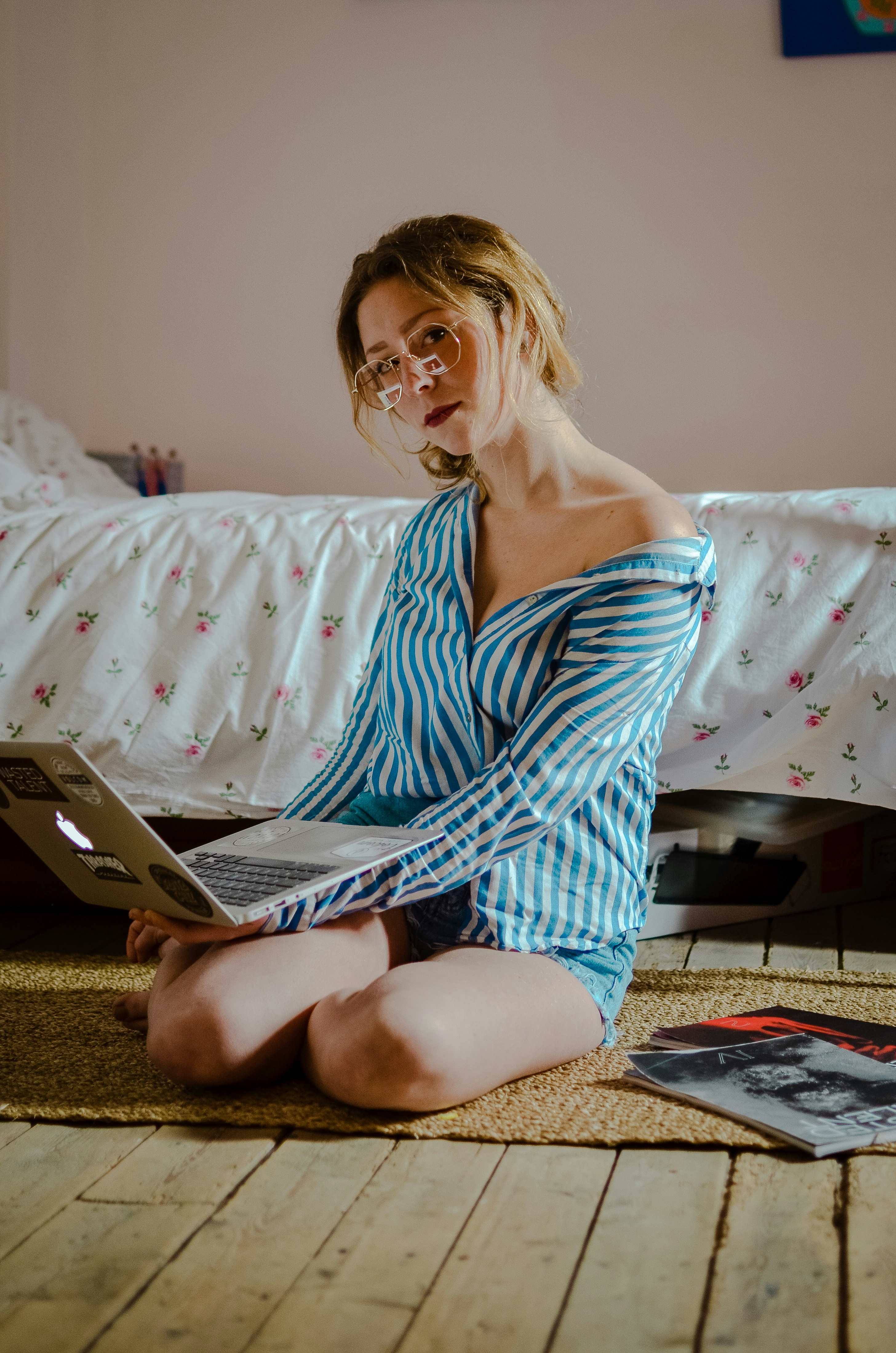Person sitting on a rug in a bedroom, using a laptop with magazines nearby.