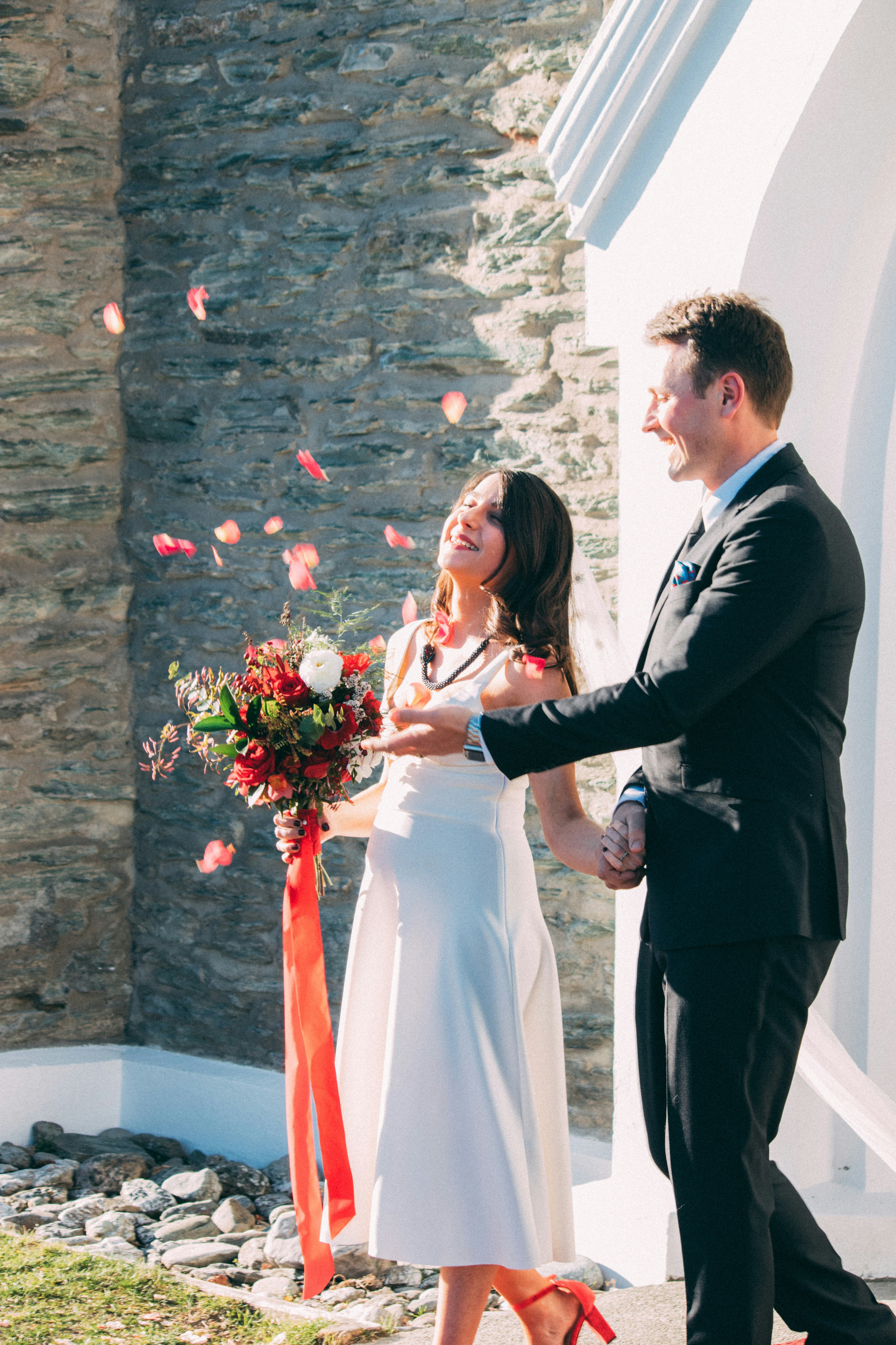 Bride and groom celebrate with a bouquet as rose petals cascade around them, capturing a joyful wedding moment.