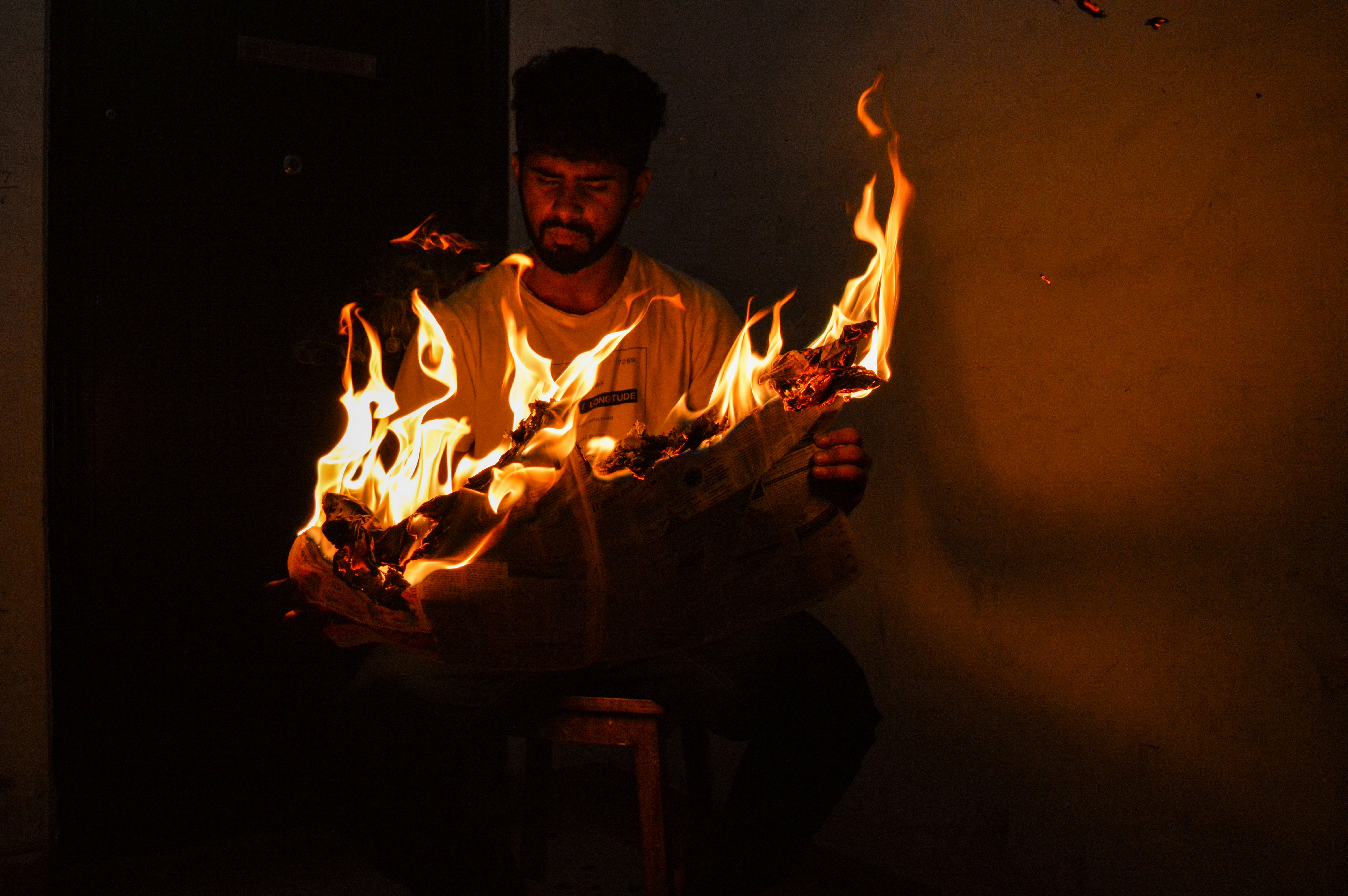 A young man sits on a stool, holding a burning piece of paper, surrounded by flickering flames in a dimly lit room.