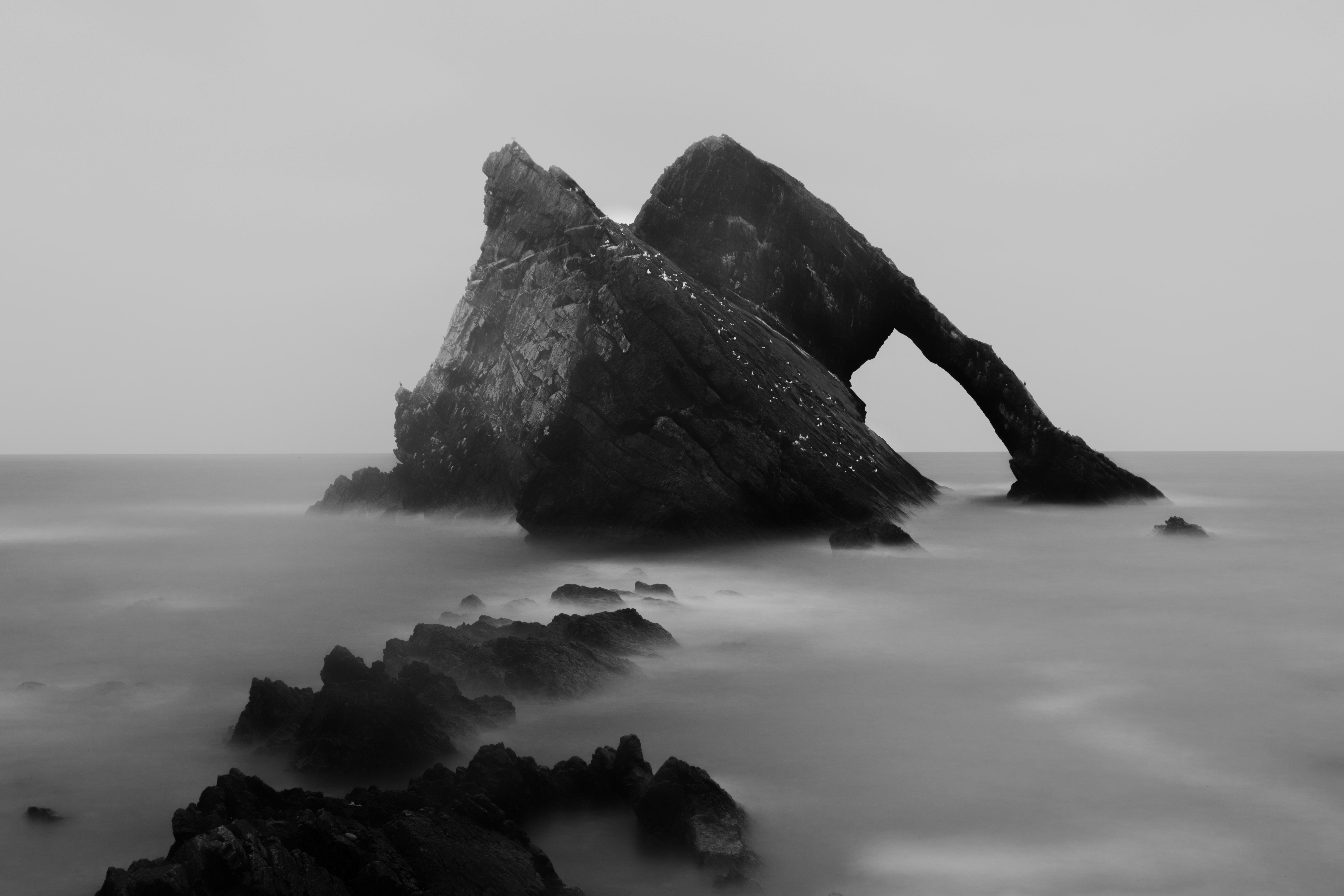 Monochrome seascape featuring a dramatic rock formation with an arch, surrounded by gentle waves and misty atmosphere.