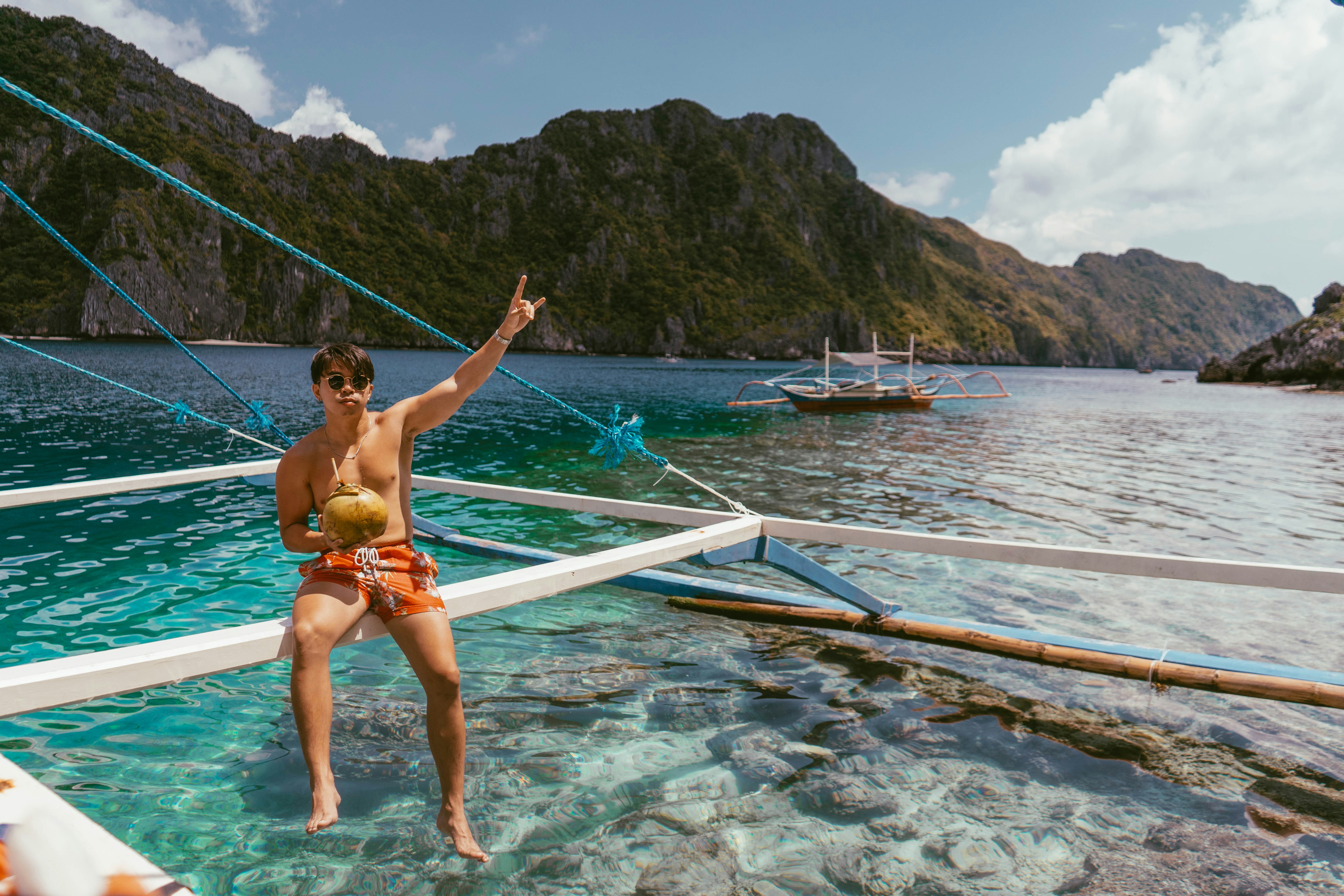 Person sitting on a boat outrigger over clear turquoise waters, holding a coconut with lush mountains in the background.