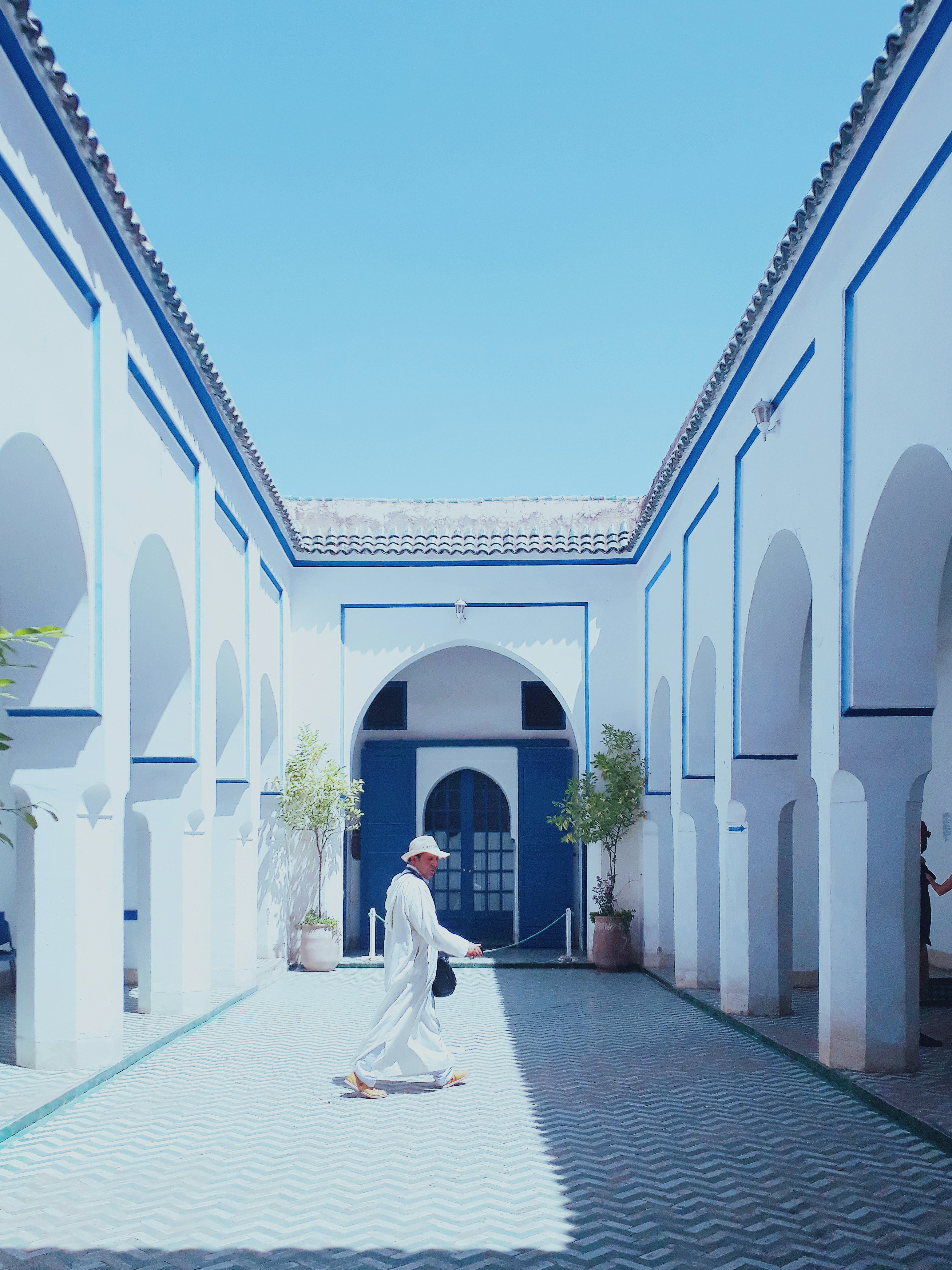 A person in traditional attire walks through a sunlit courtyard adorned with blue and white architecture, framed by lush greenery. The scene captures a moment of tranquility.