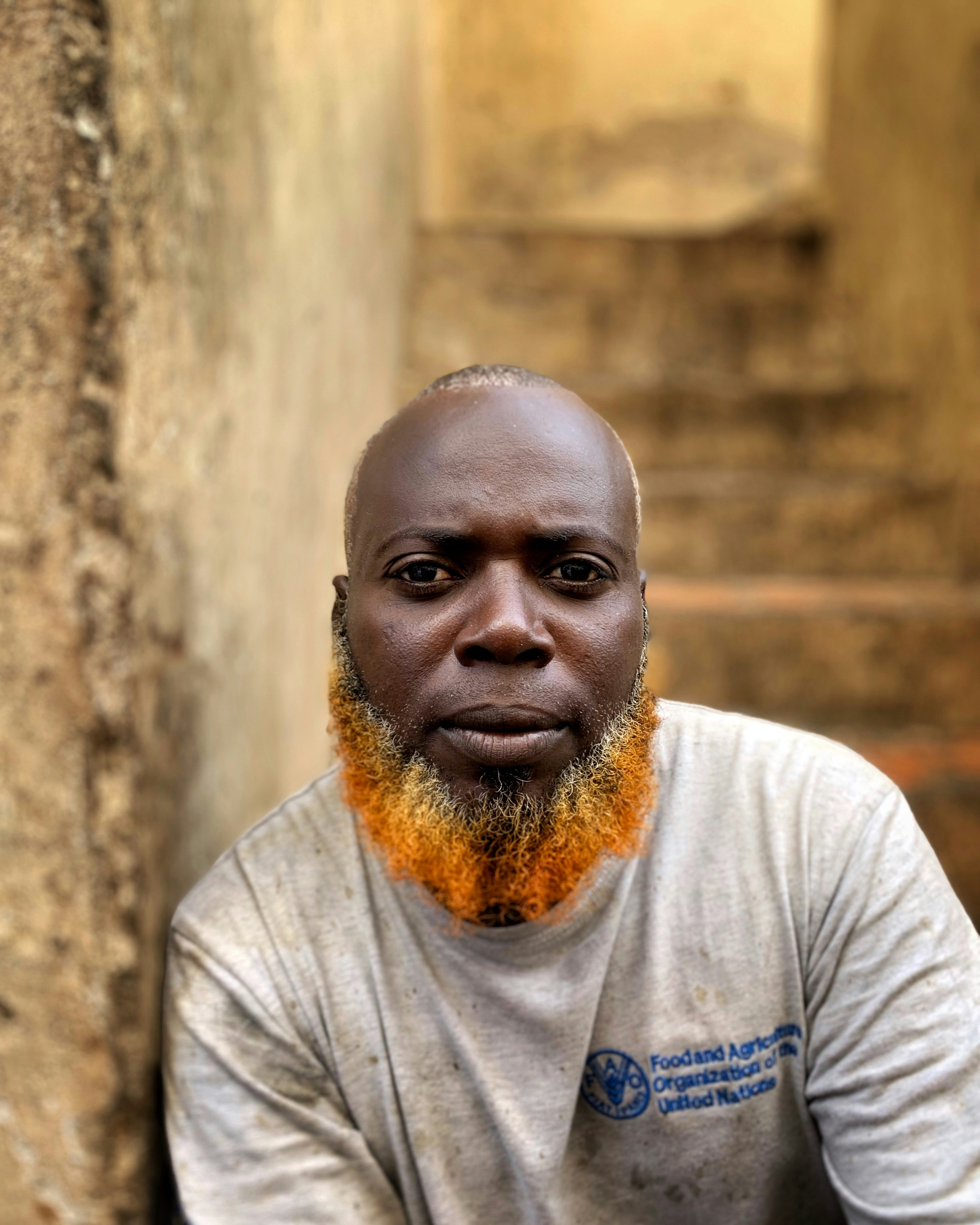 A man with a striking orange beard sits against a textured wall, exuding a sense of calm amidst urban surroundings.