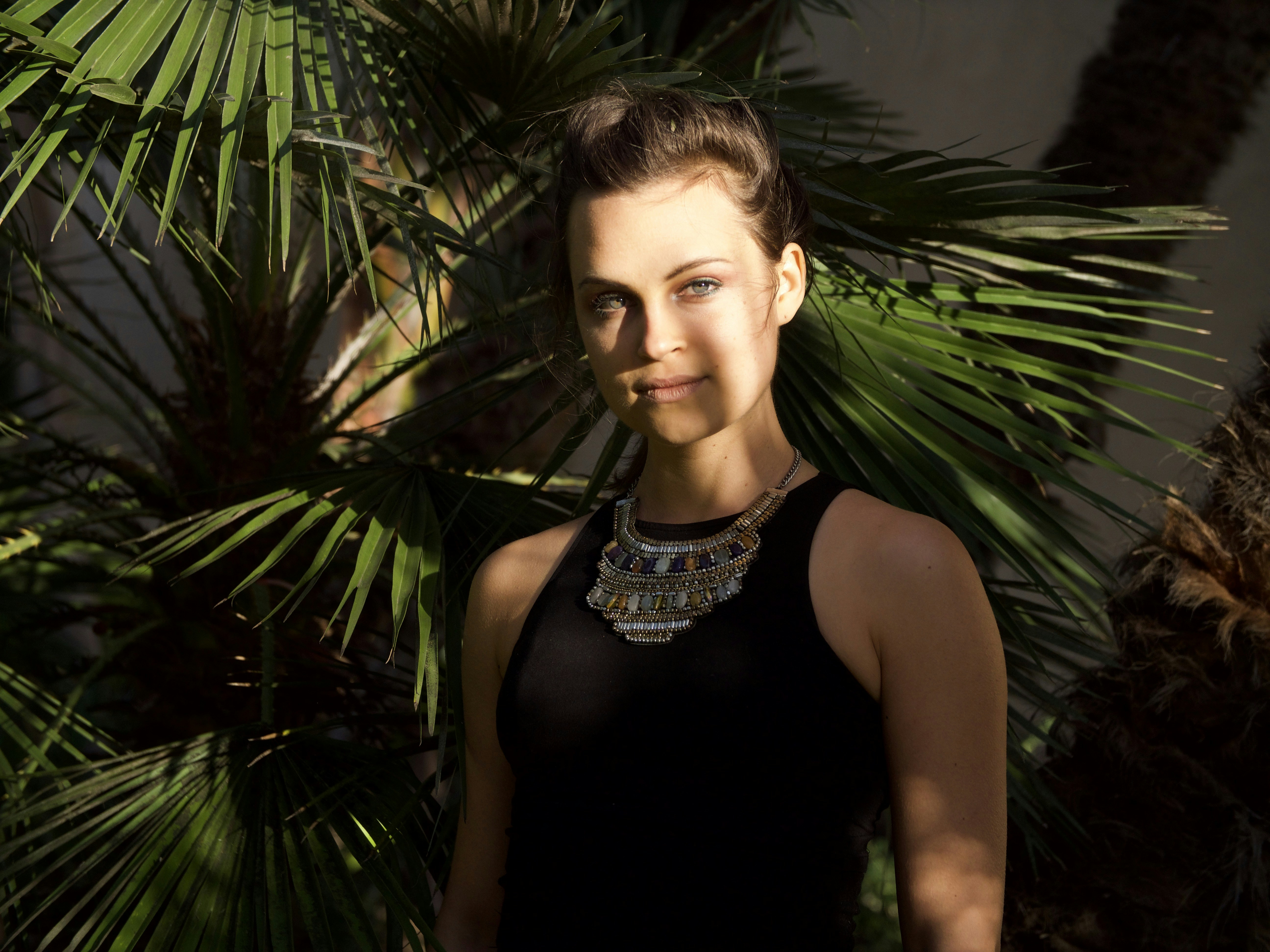 Young woman with a striking necklace stands amidst lush palm leaves, illuminated by soft sunlight filtering through the foliage.