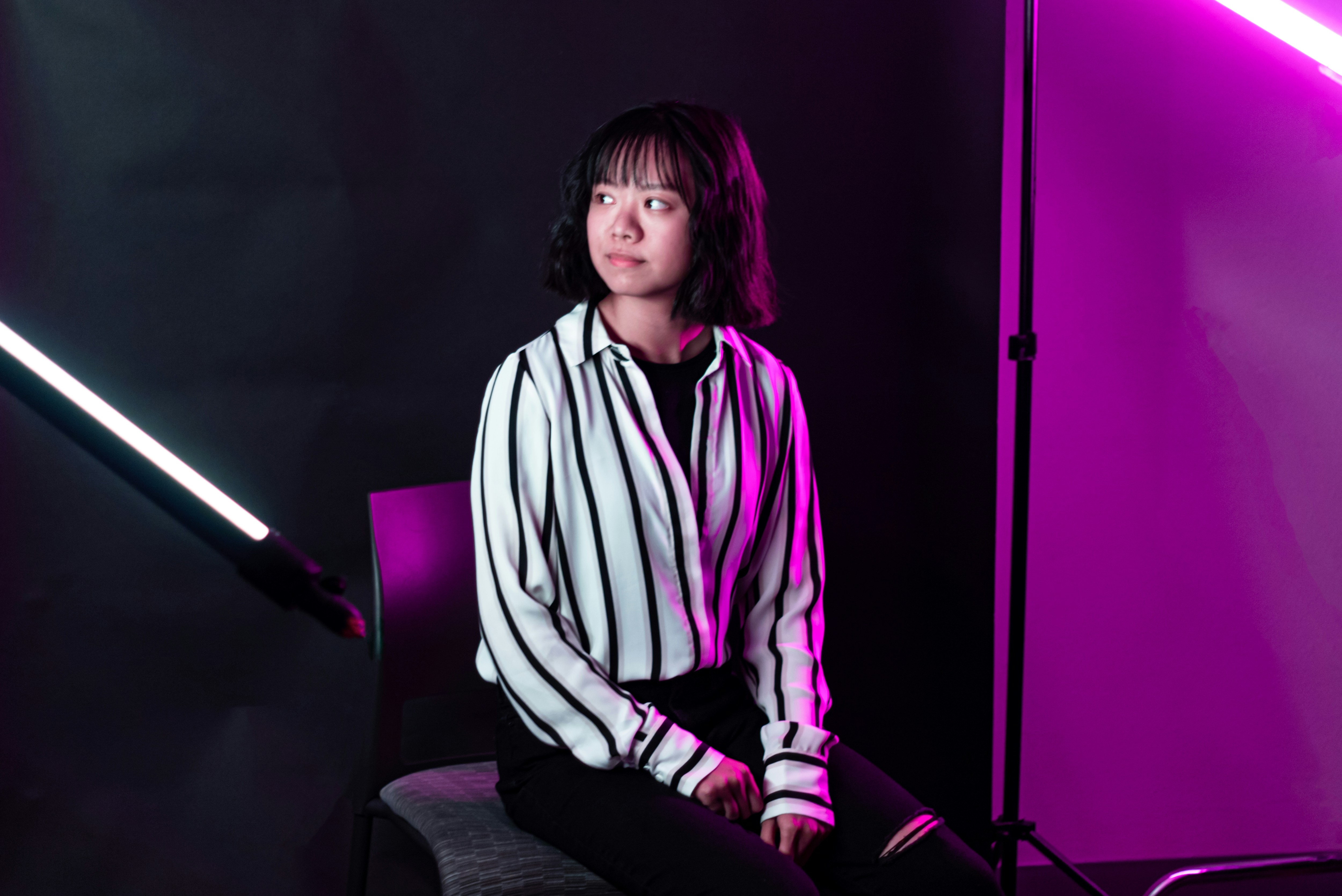 A young woman seated against a dark backdrop, illuminated by vibrant neon lights, showcasing a modern fashion aesthetic.
