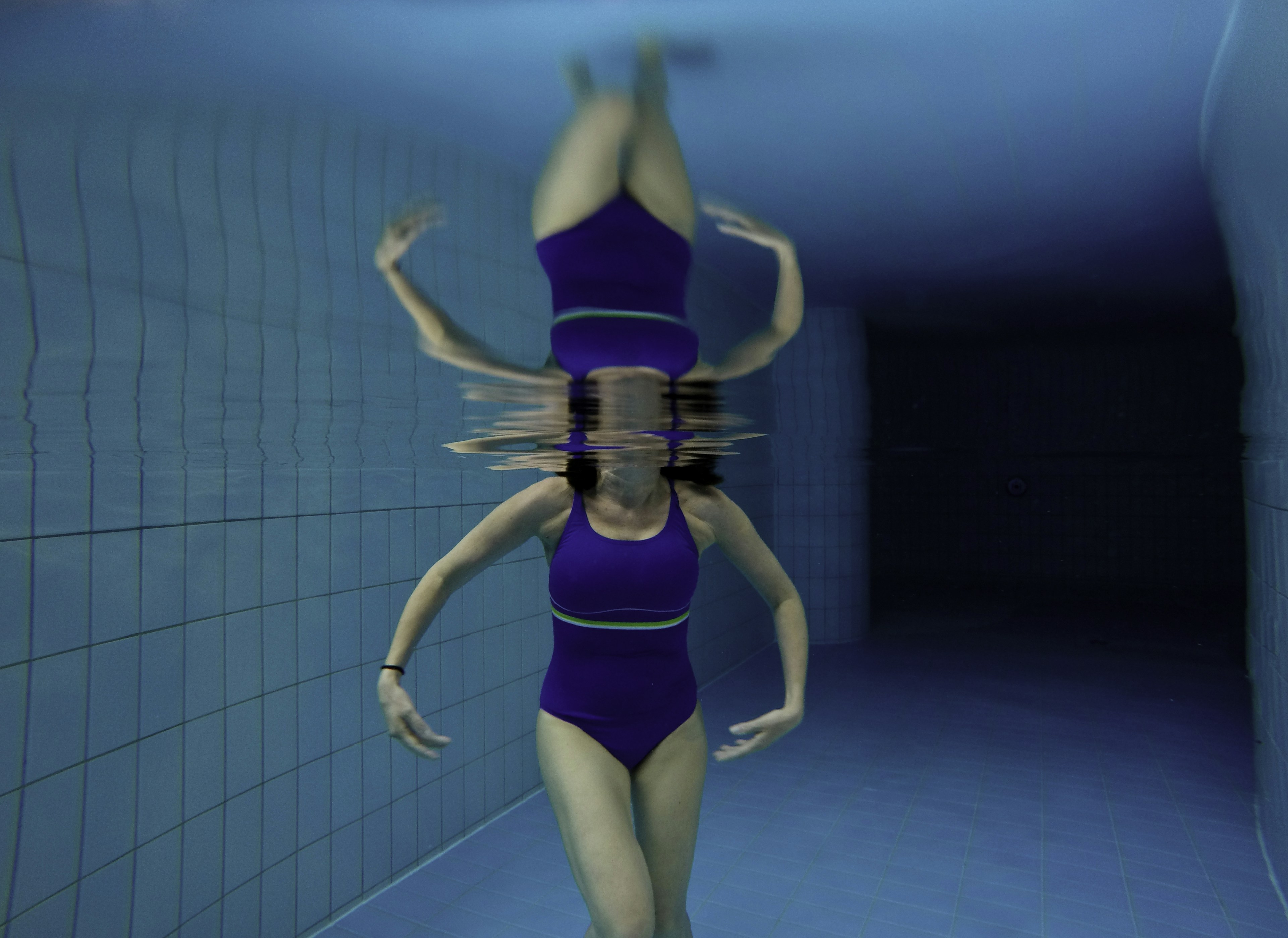 Woman in a blue bodysuit performing a graceful underwater pose in a pool, creating a mirrored reflection.