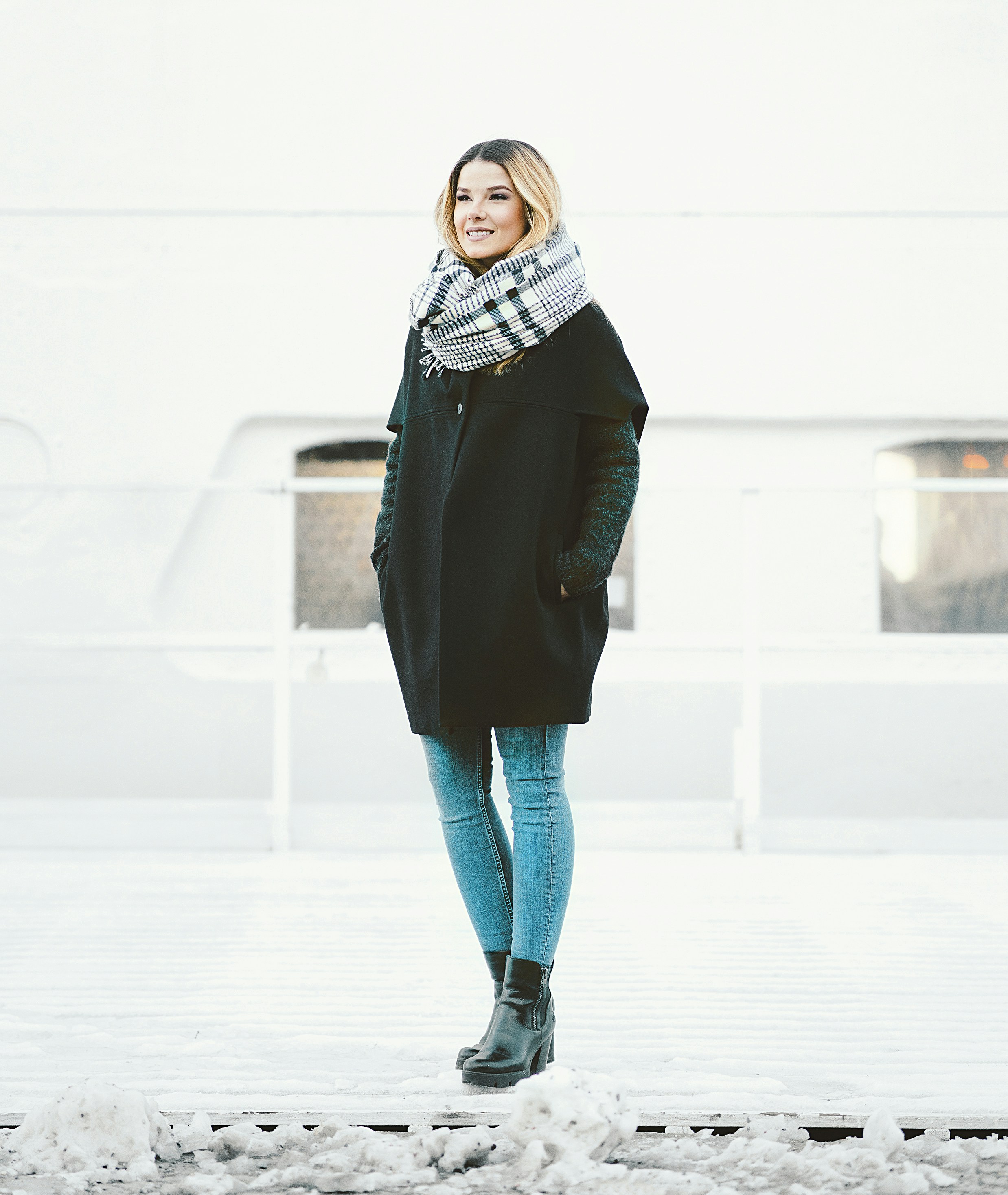 Woman in a stylish black coat and plaid scarf stands confidently on a snowy urban street, exuding warmth and fashion. Her pose contrasts with the minimalist background.