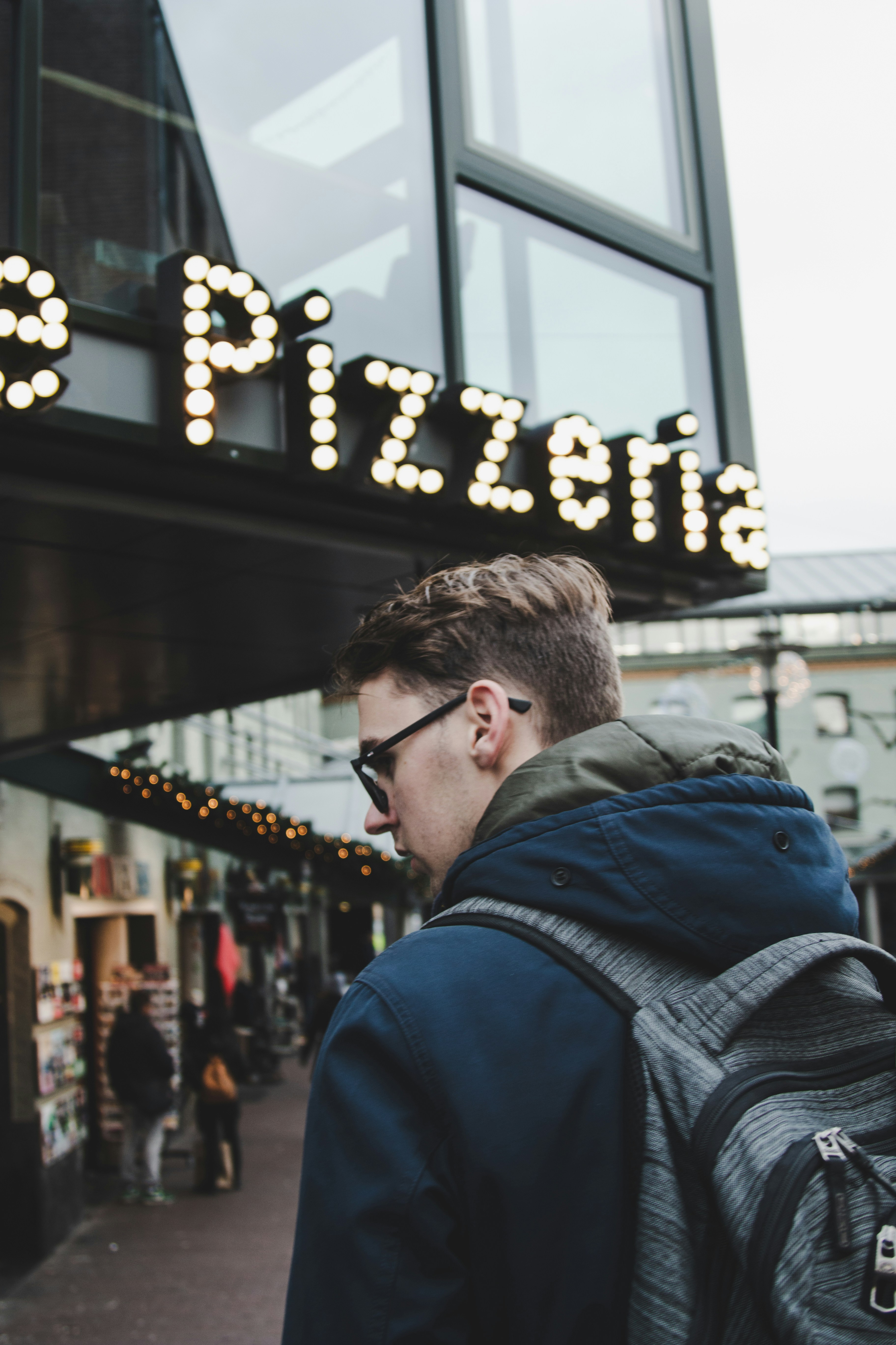 A young man in sunglasses walks past a brightly lit pizzeria sign in an urban setting, capturing the essence of city life. The ambiance reflects a lively atmosphere.