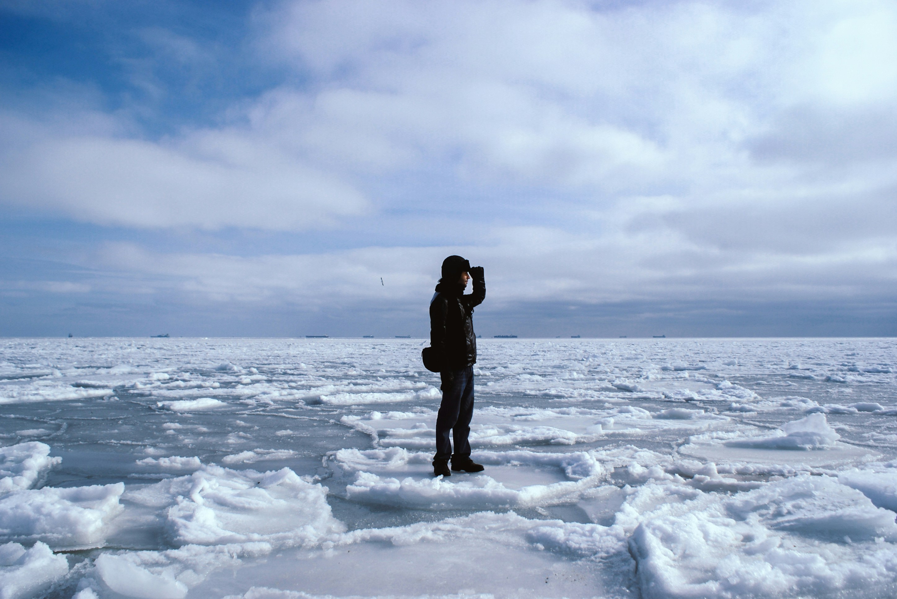 Person standing on a frozen sea, surrounded by broken ice under a vast, cloudy sky.