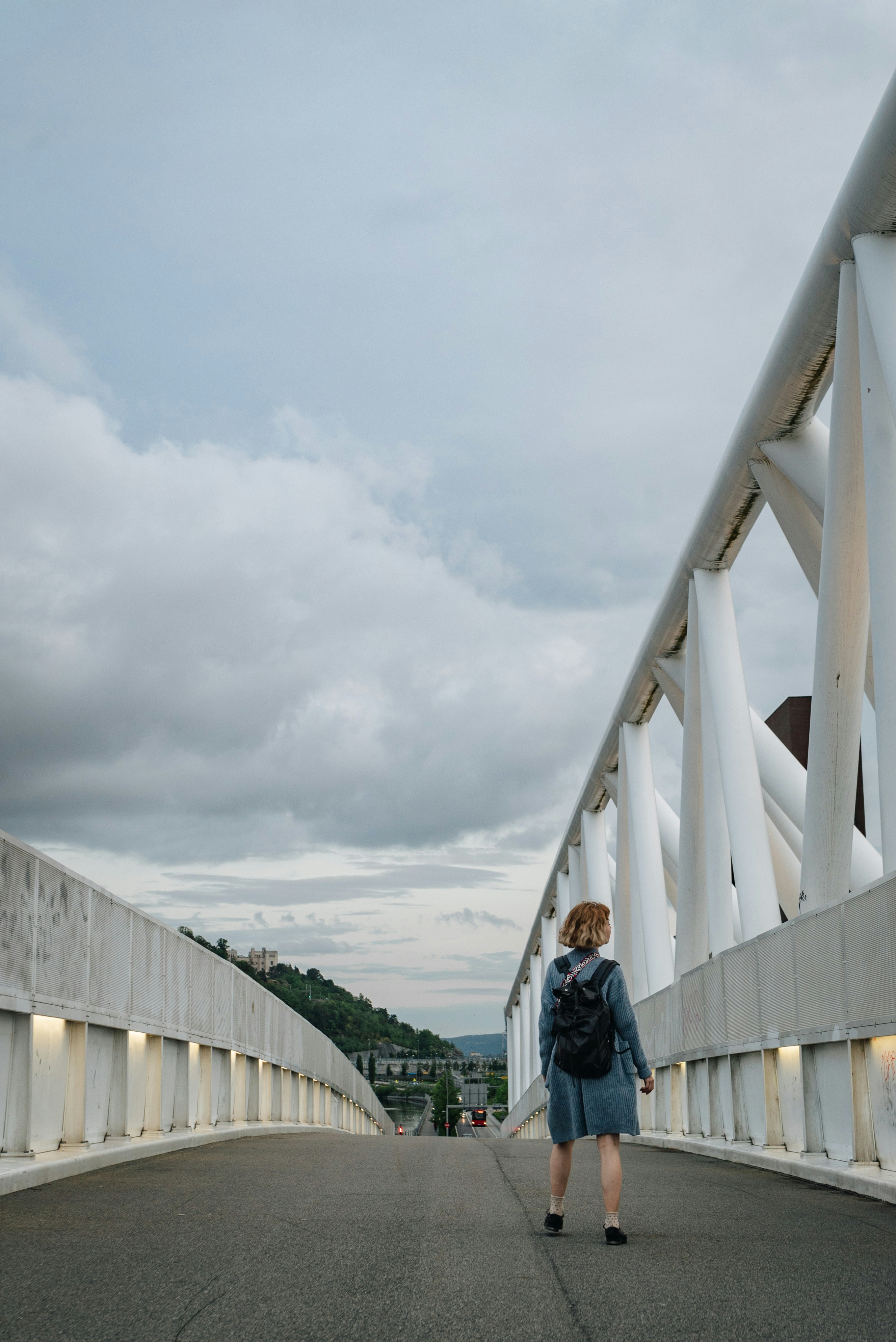 A lone figure walks along a contemporary bridge, flanked by striking white supports under a cloudy sky.