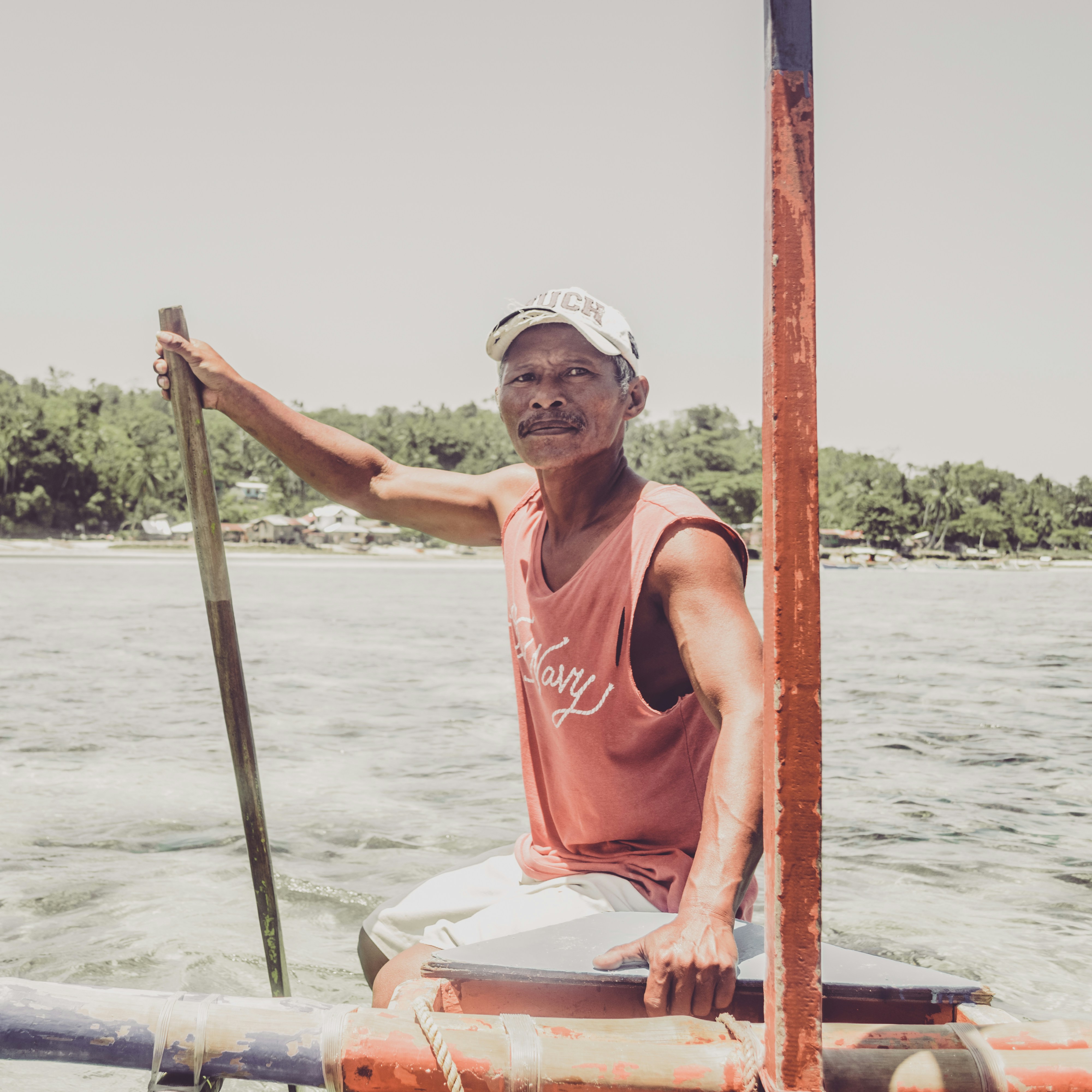 A fisherman in a red tank top paddles a small boat in calm waters, surrounded by lush greenery and distant shoreline.