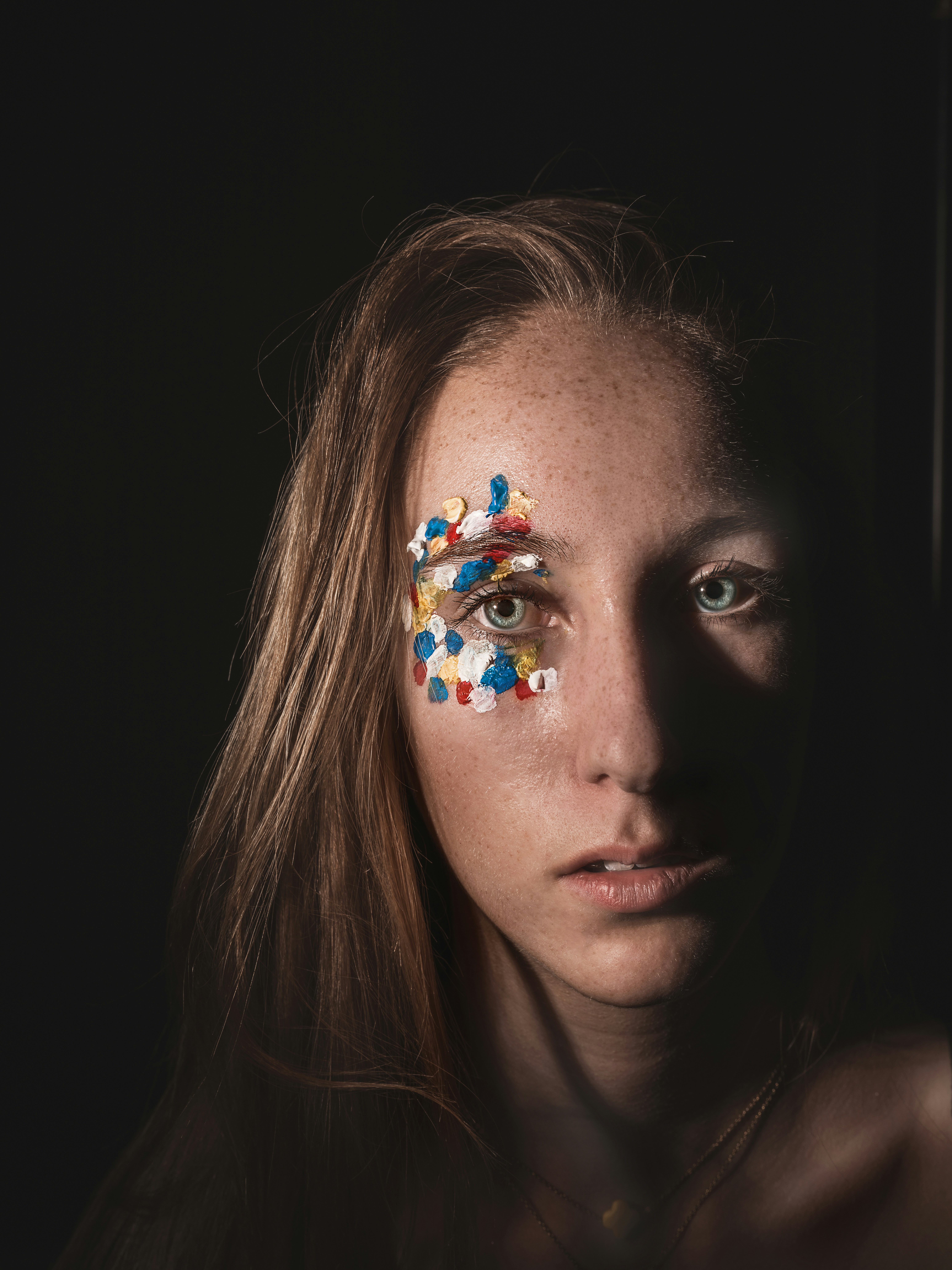 Portrait of a young woman with colorful paint flakes on one eye, lit from the side to reveal freckles and a soft, moody expression. The composition centers the face against a dark background, highlighting the eye and texture.