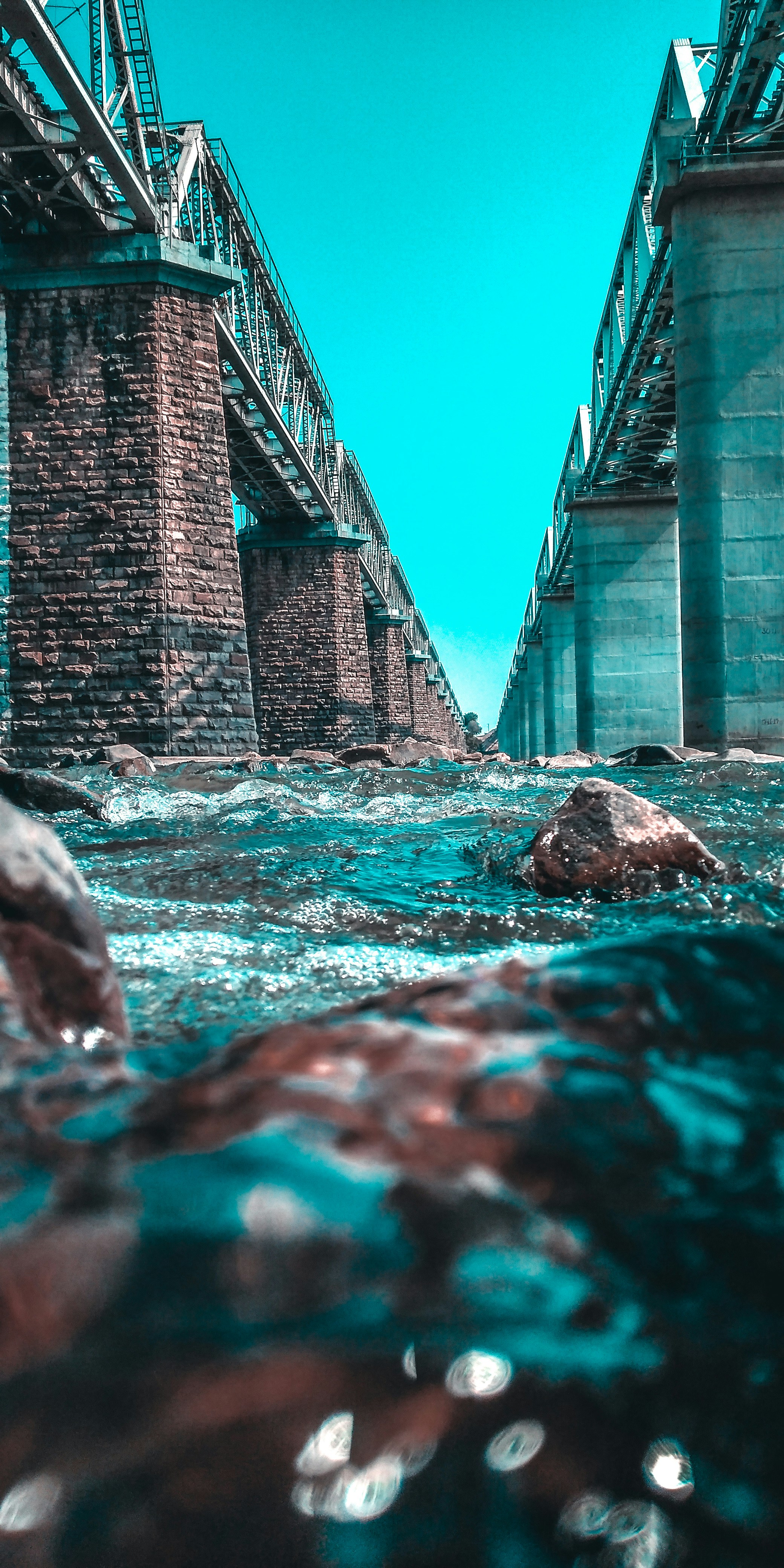 View from beneath a bridge, showcasing the flowing river and stone pillars. The vibrant turquoise water contrasts with the rugged bridge structure.
