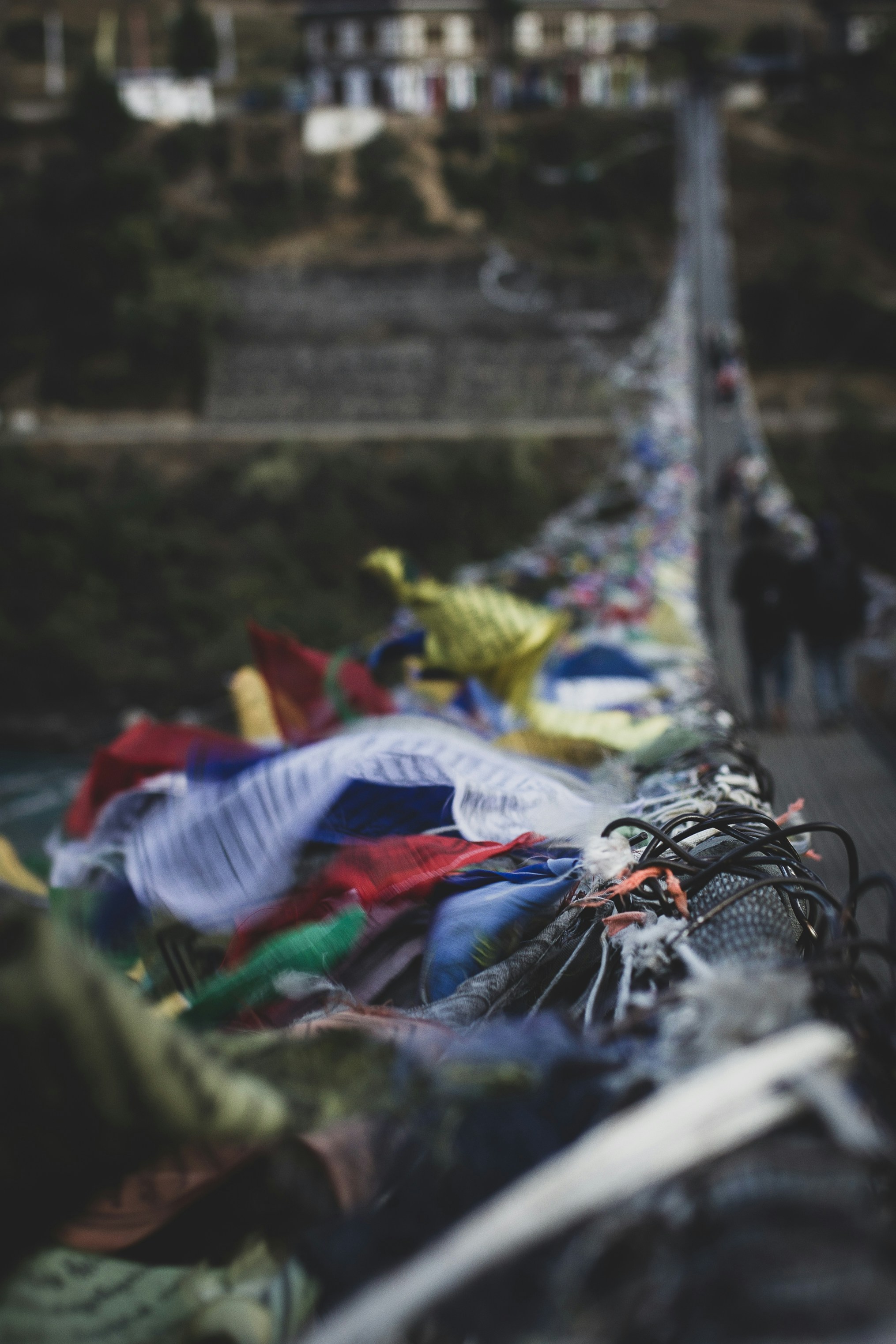 Colorful prayer flags fluttering on a suspension bridge, with a blurred background of distant buildings and nature. The scene captures the essence of cultural reverence and natural beauty.
