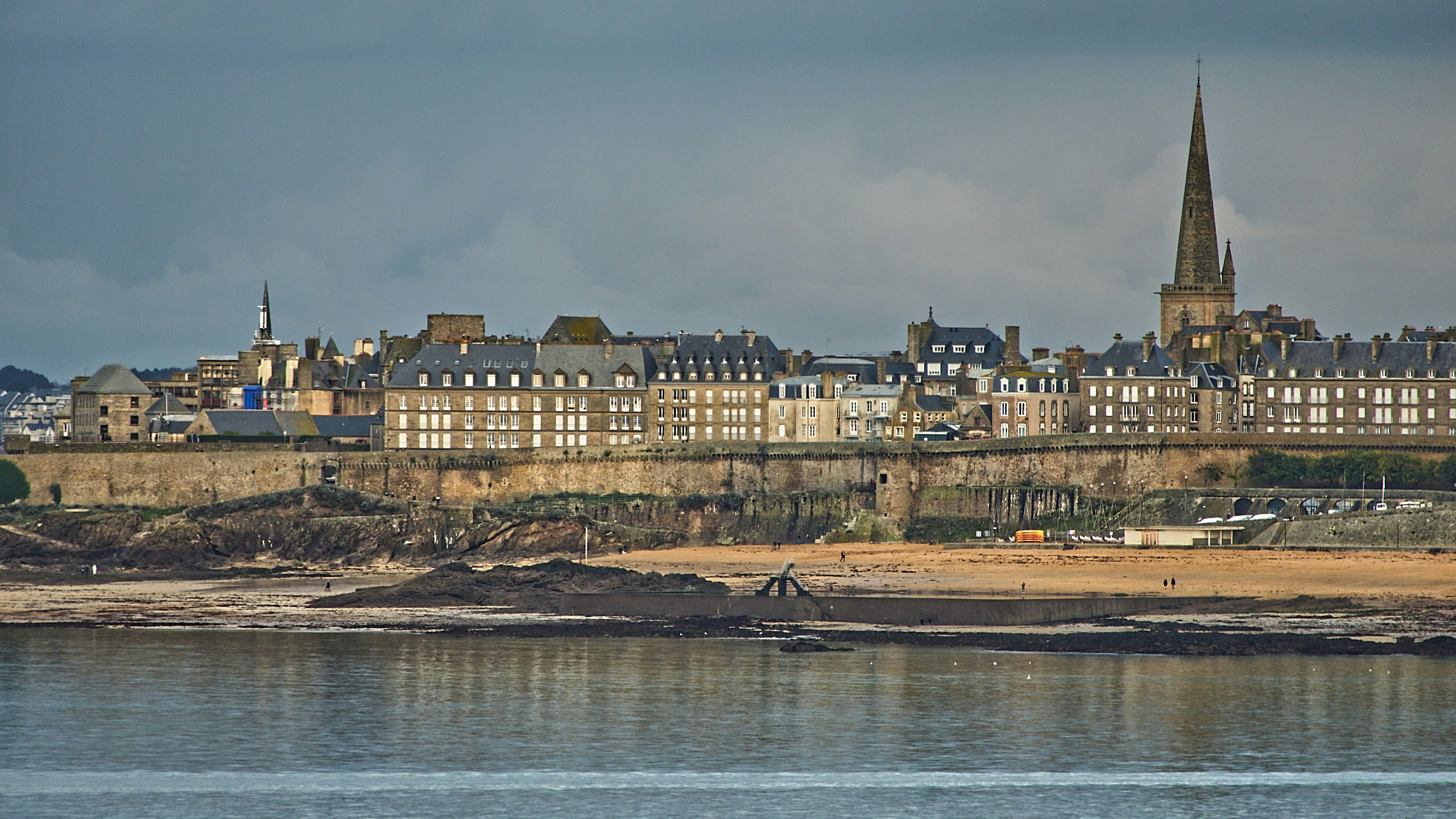 Historic buildings and a tall church spire of Saint-Malo under a cloudy sky, viewed from across the water.