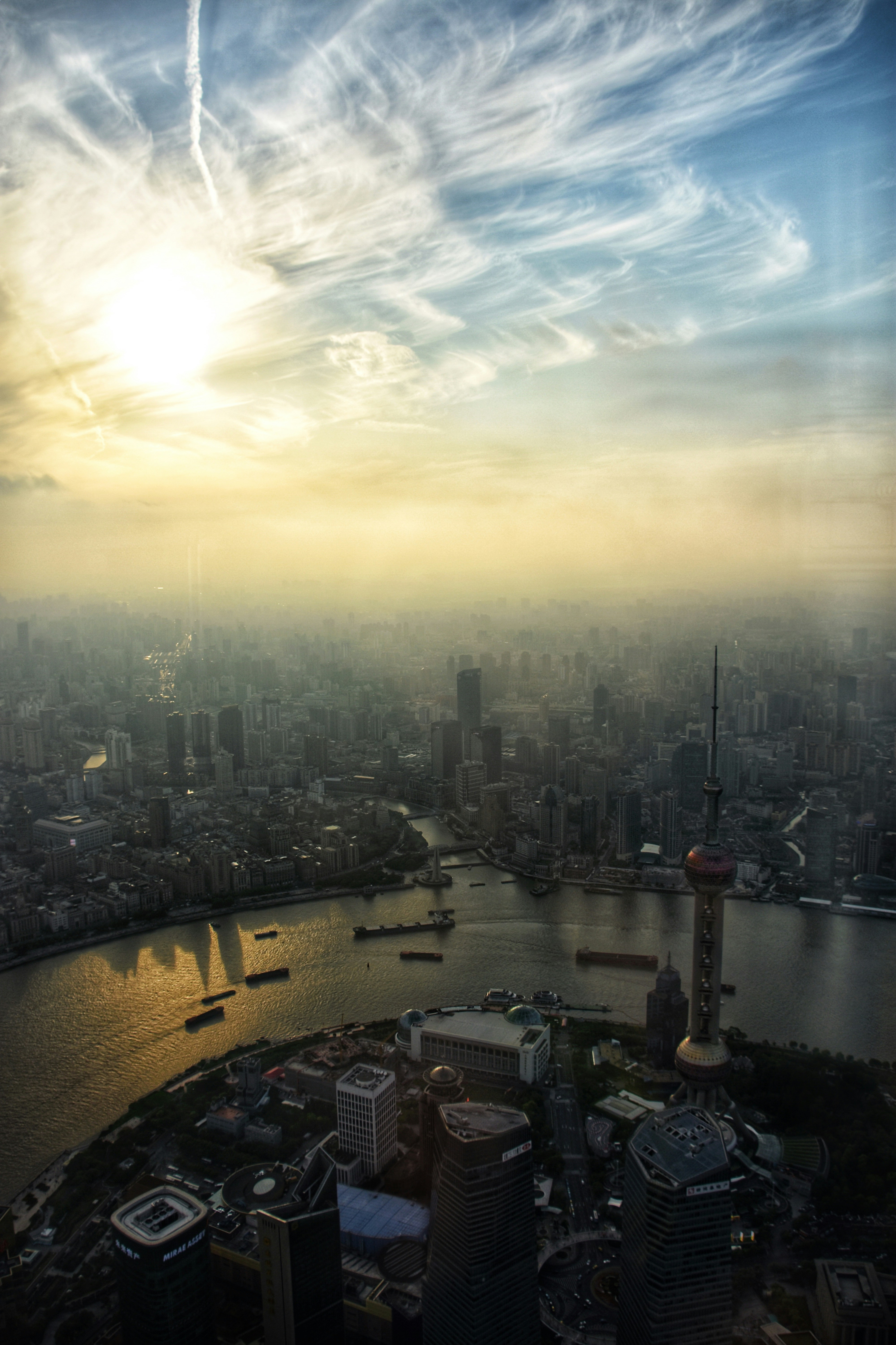 Aerial view of Shanghai's skyline shrouded in haze, featuring the iconic Oriental Pearl Tower and the winding Huangpu River reflecting the sunset hues.