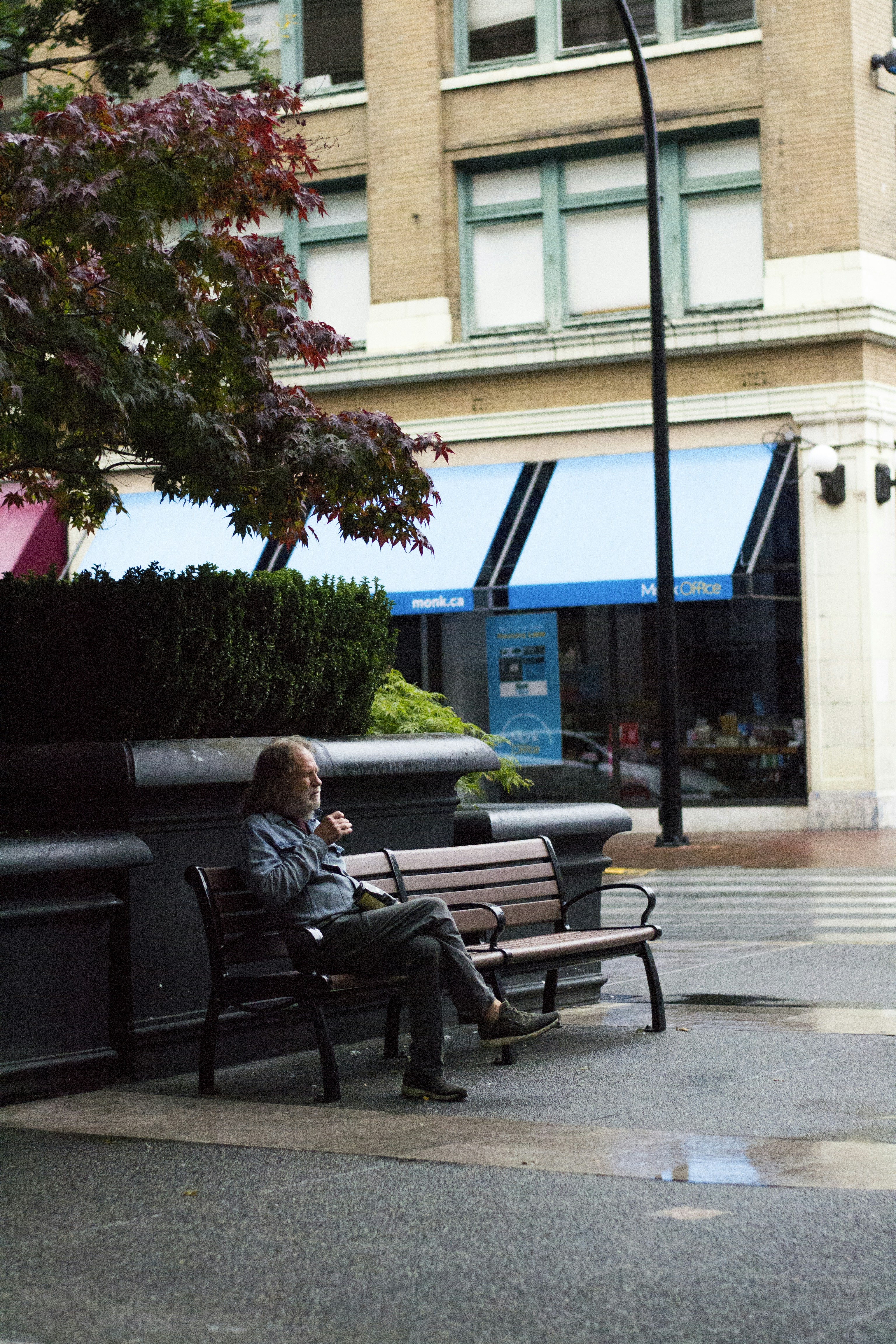 An elderly man sits quietly on a bench, surrounded by greenery and urban architecture, lost in thought. The scene captures a peaceful pause in a bustling city.