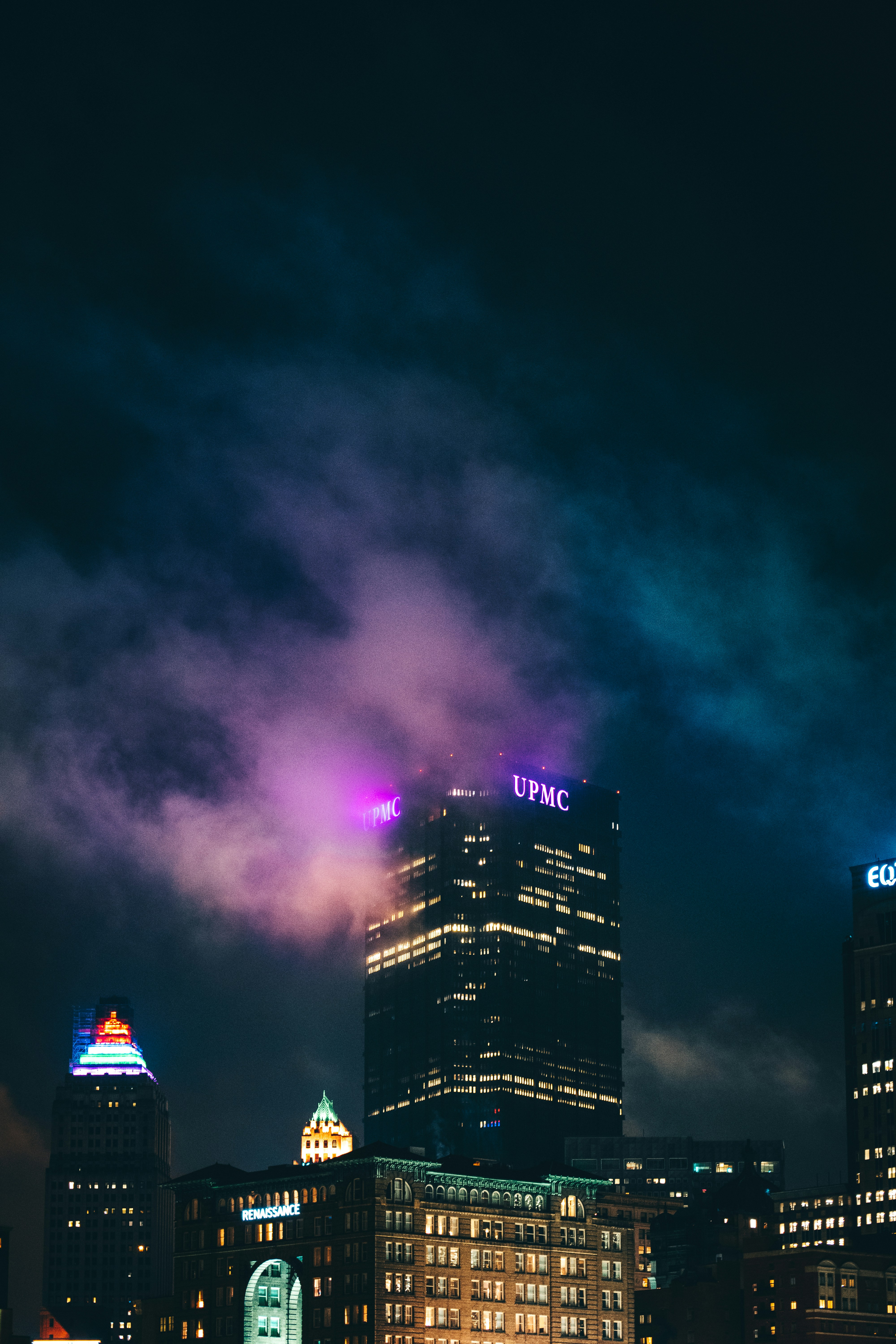 Illuminated skyscrapers rise against a moody night sky, with clouds swirling around the UPMC building. The scene captures the essence of a vibrant cityscape at dusk.