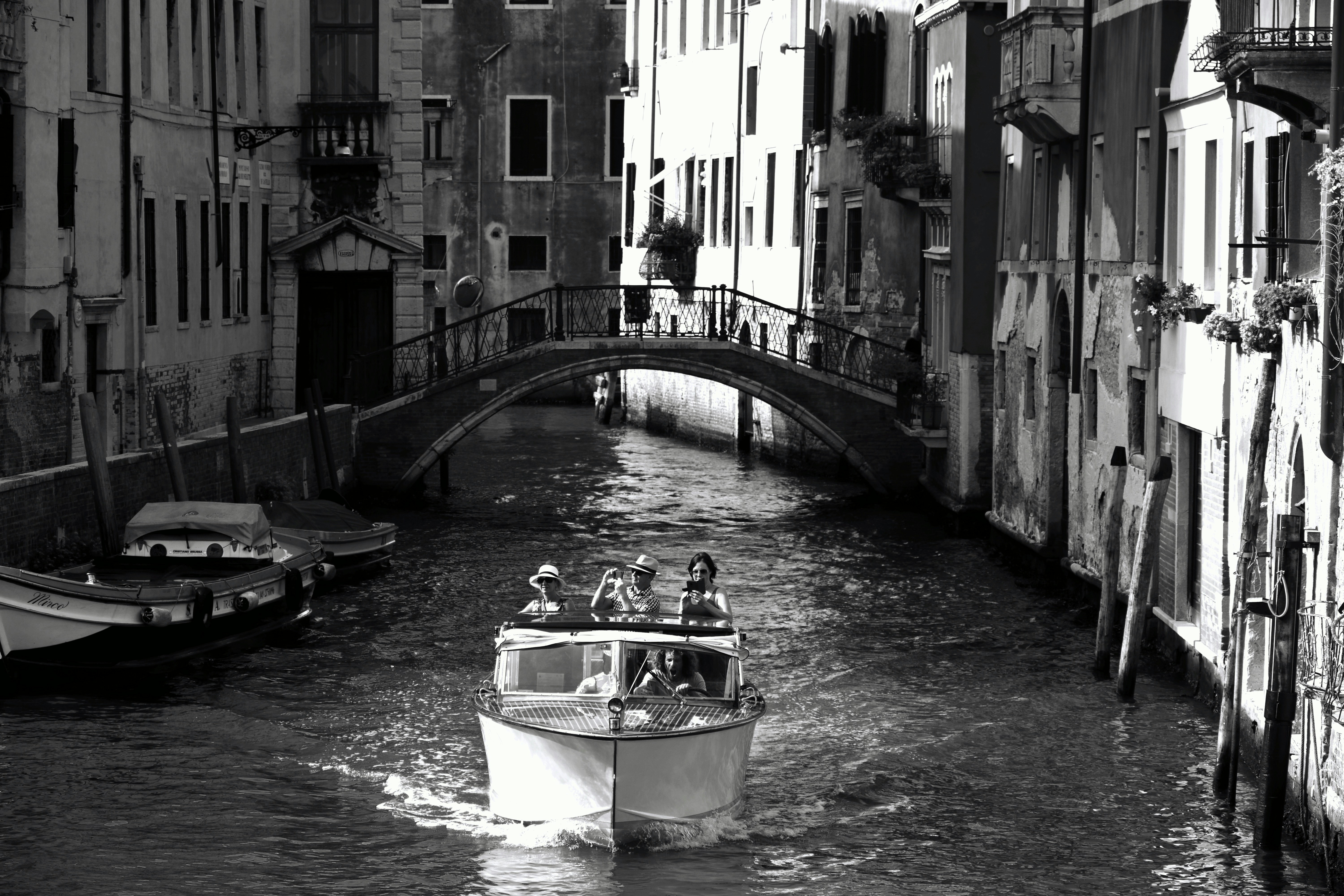 A motorboat glides through a narrow canal in Venice, flanked by historic buildings and a quaint bridge. The scene is rendered in black and white, emphasizing the timeless charm of the city.