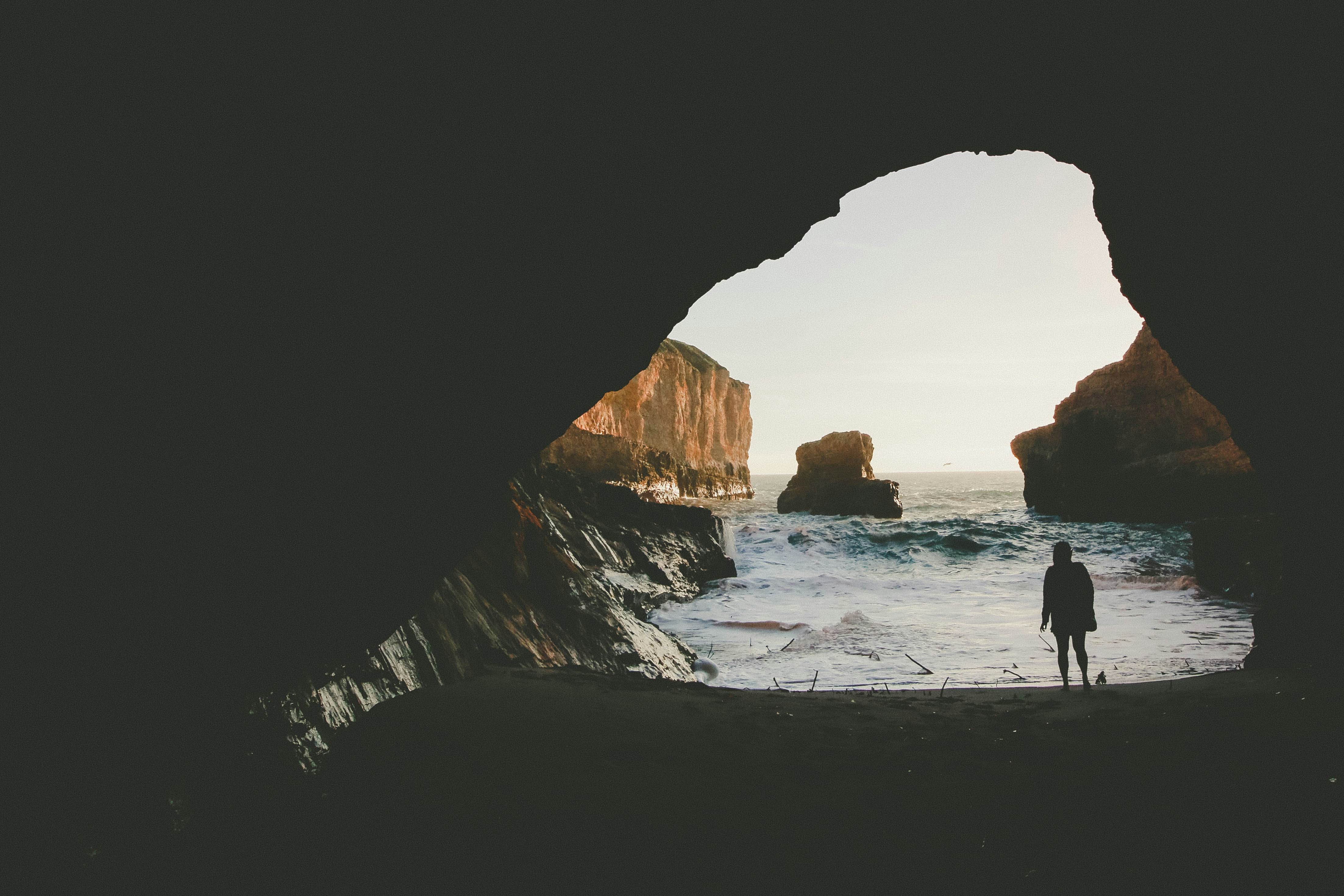 Silhouette of a person standing at the entrance of an ocean cave, overlooking turbulent waves and rocky cliffs at sunset.