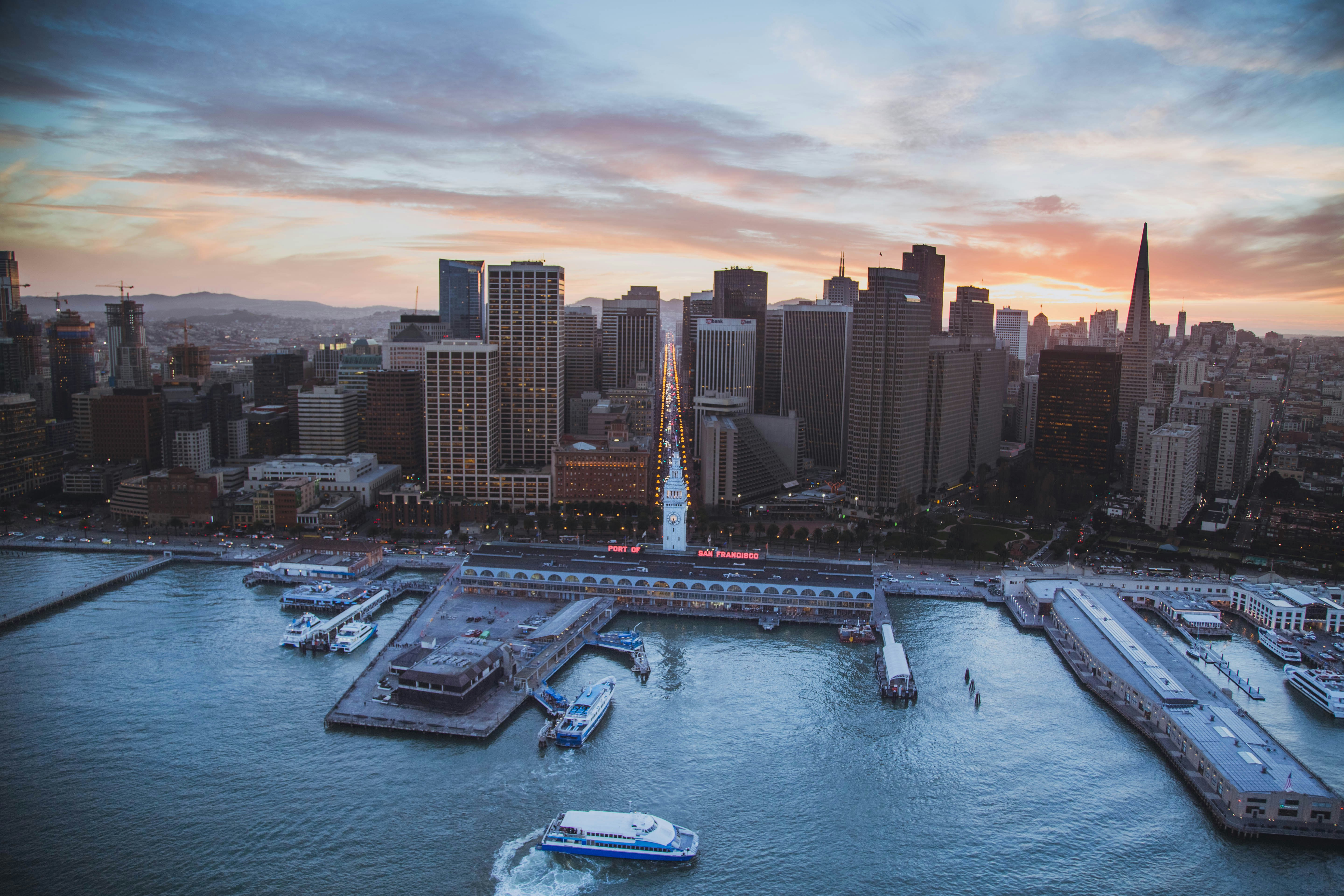 Aerial view of a city skyline at sunset with a waterfront and boats in the foreground.