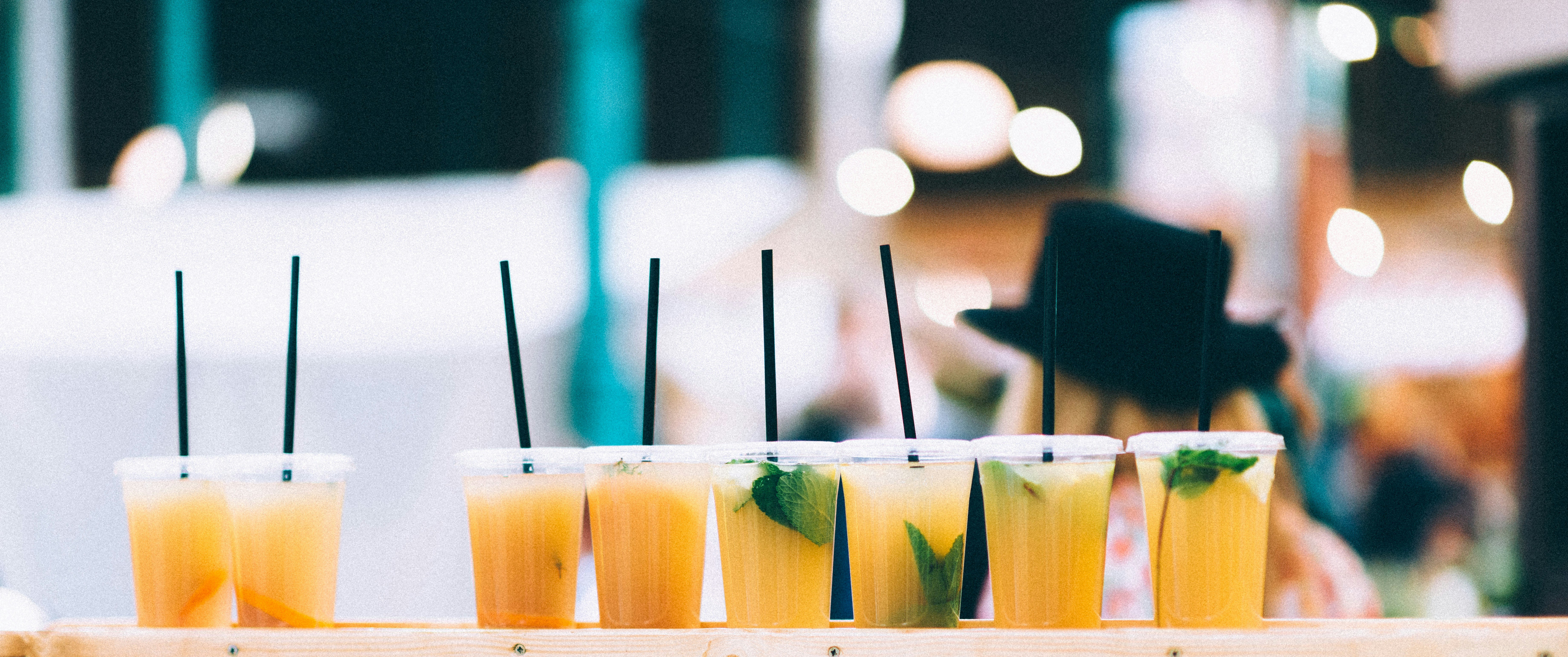 Lineup of vibrant orange drinks garnished with mint, set against a blurred market backdrop. The scene conveys a lively atmosphere.