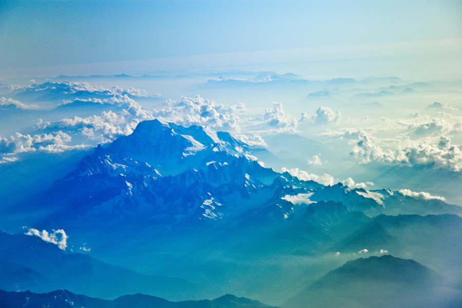 Snow-dusted mountain peak emerging through layers of blue mist and clouds, with distant ridgelines fading into a pale sky.