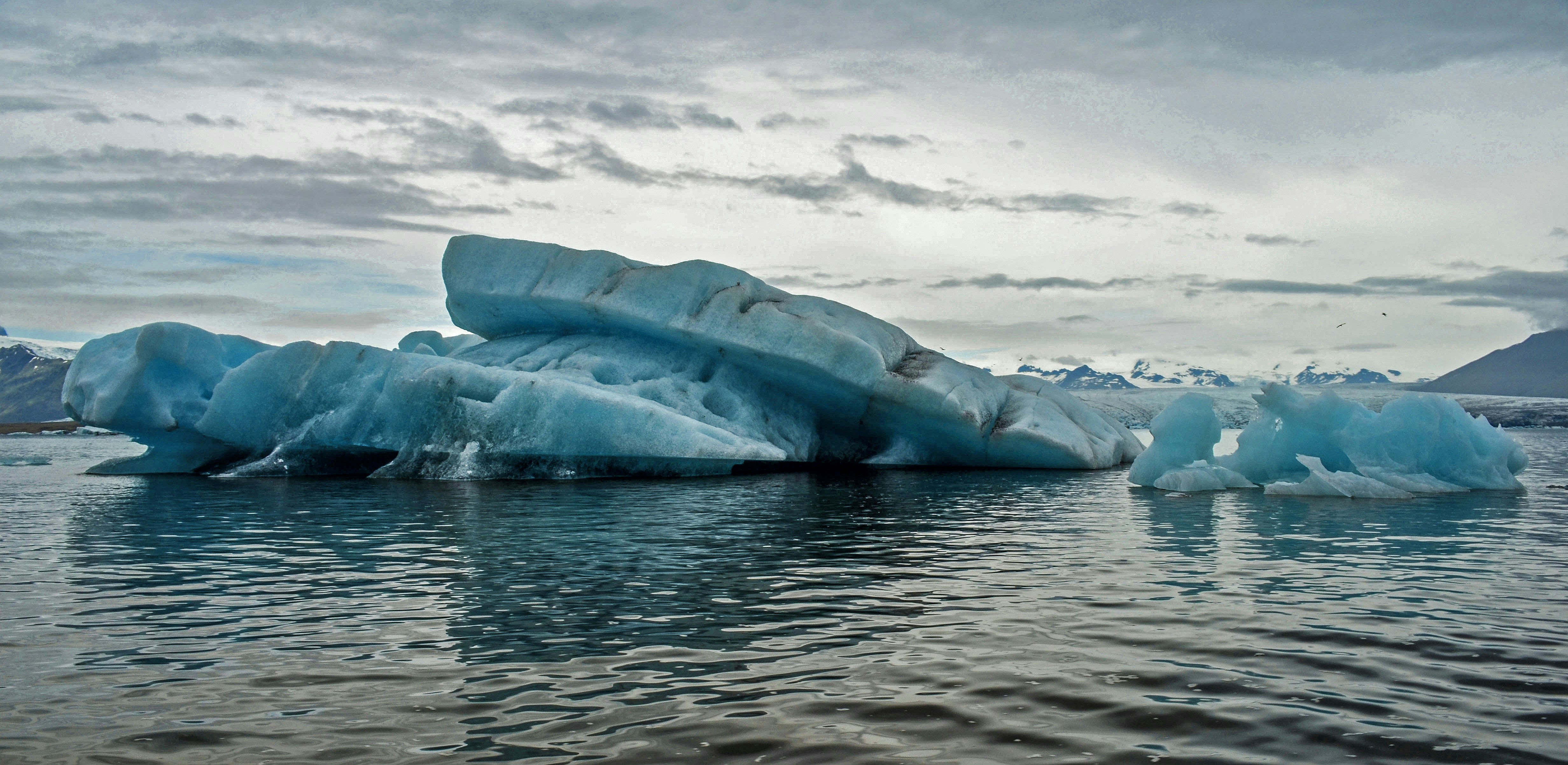 A majestic iceberg floats serenely in calm waters, showcasing its intricate blue hues and layered structure against a cloudy sky.
