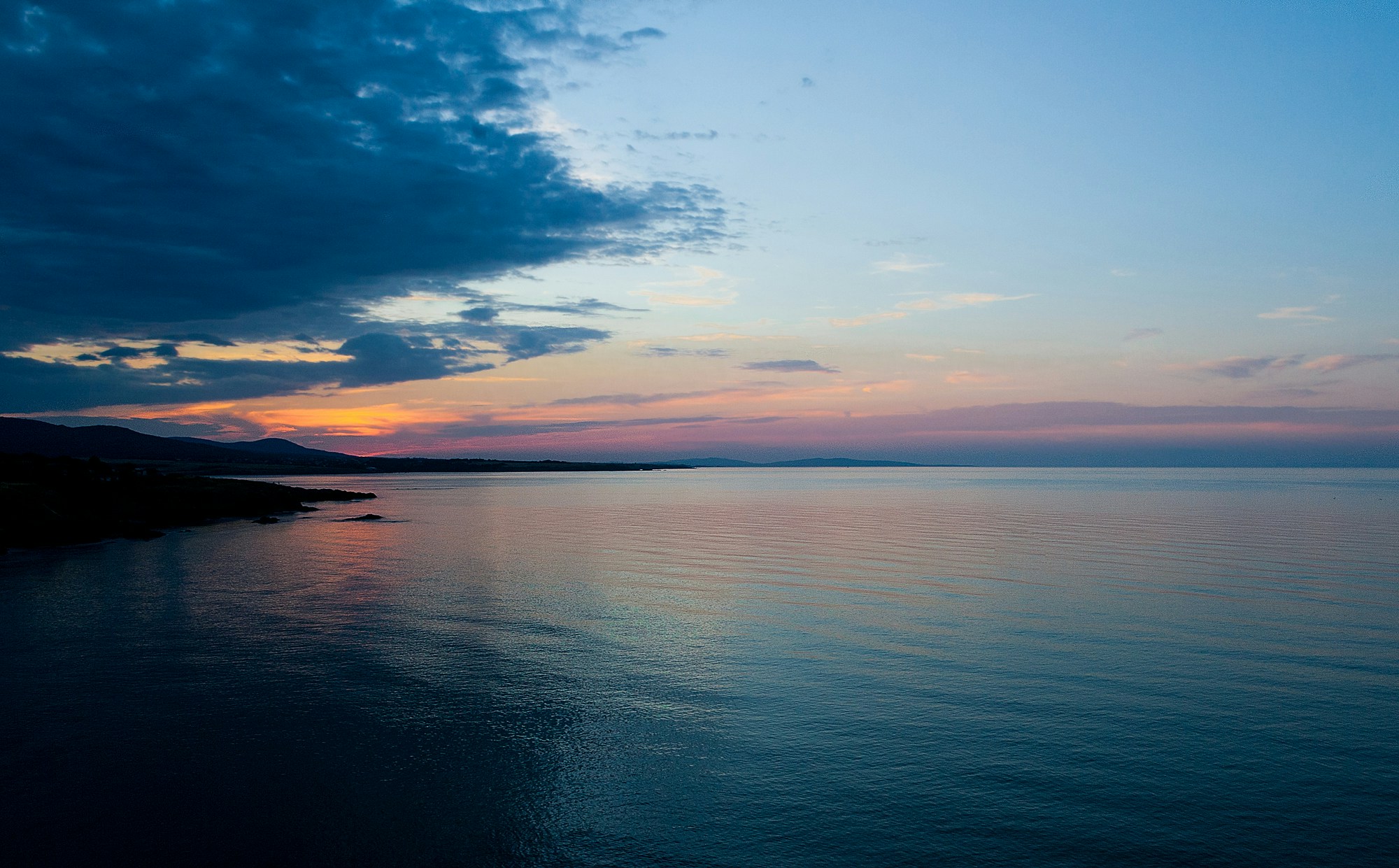 Calm sea reflecting the subtle hues of a sunset sky with silhouetted clouds and distant coastline.
