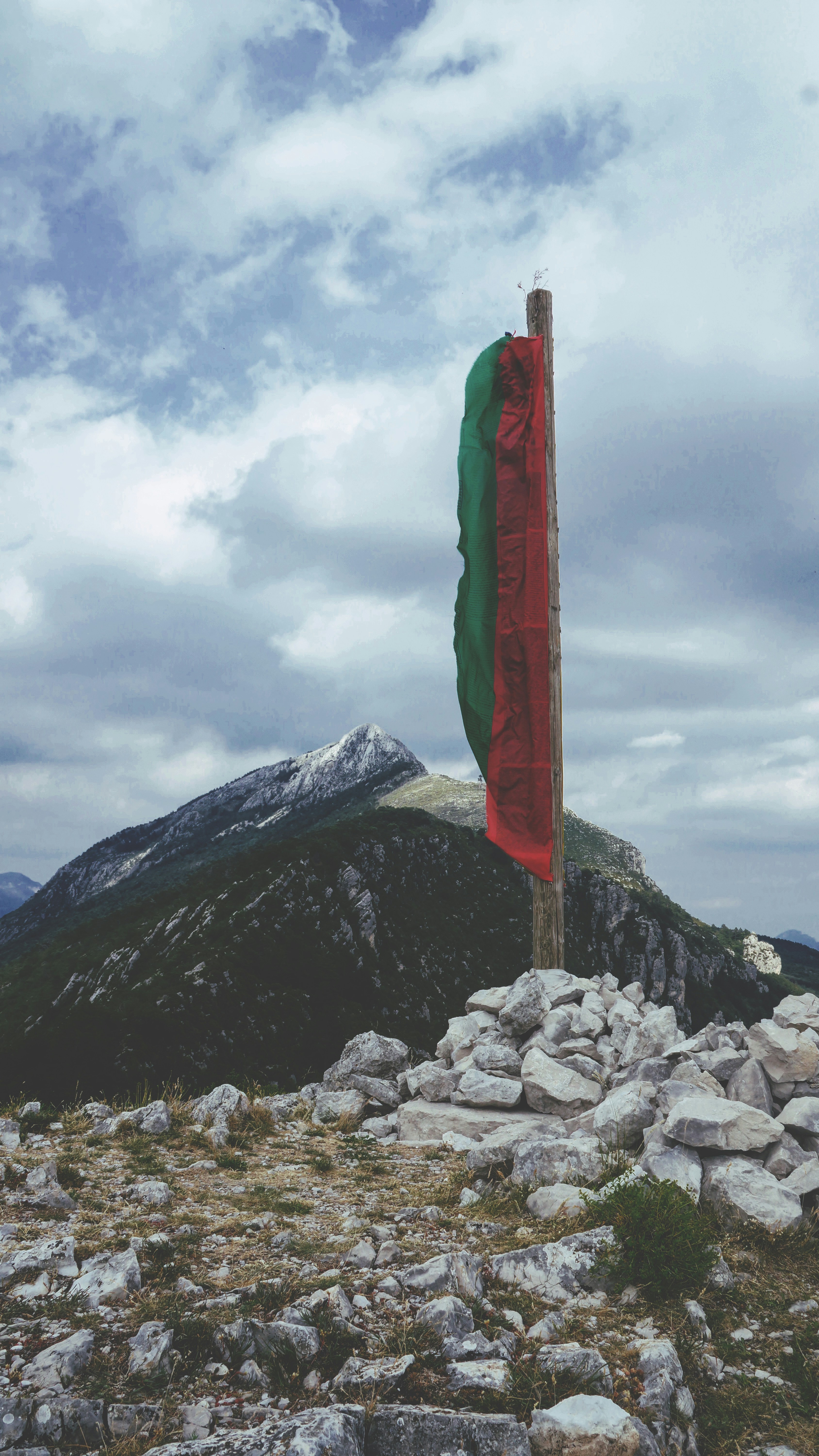 Green and red flag on a wooden pole atop a rocky mountain peak under a cloudy sky.