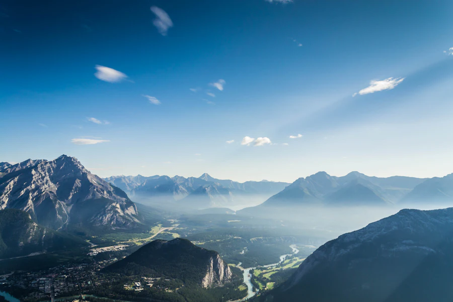 Sweeping alpine valley with rugged peaks, a winding river, and soft haze under a bright blue sky. Sunlit ridges frame the distant mountains.