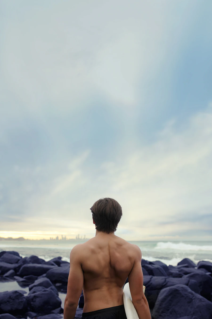 Shirtless man standing with a surfboard among dark rocks, facing the ocean under a pale, cloudy sky.