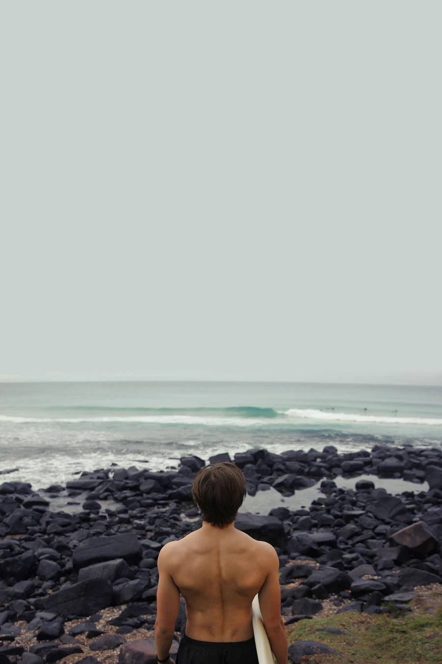 Surfer gazes out at the ocean, contemplating the surf conditions while holding a surfboard, with rocky shoreline in the foreground. 