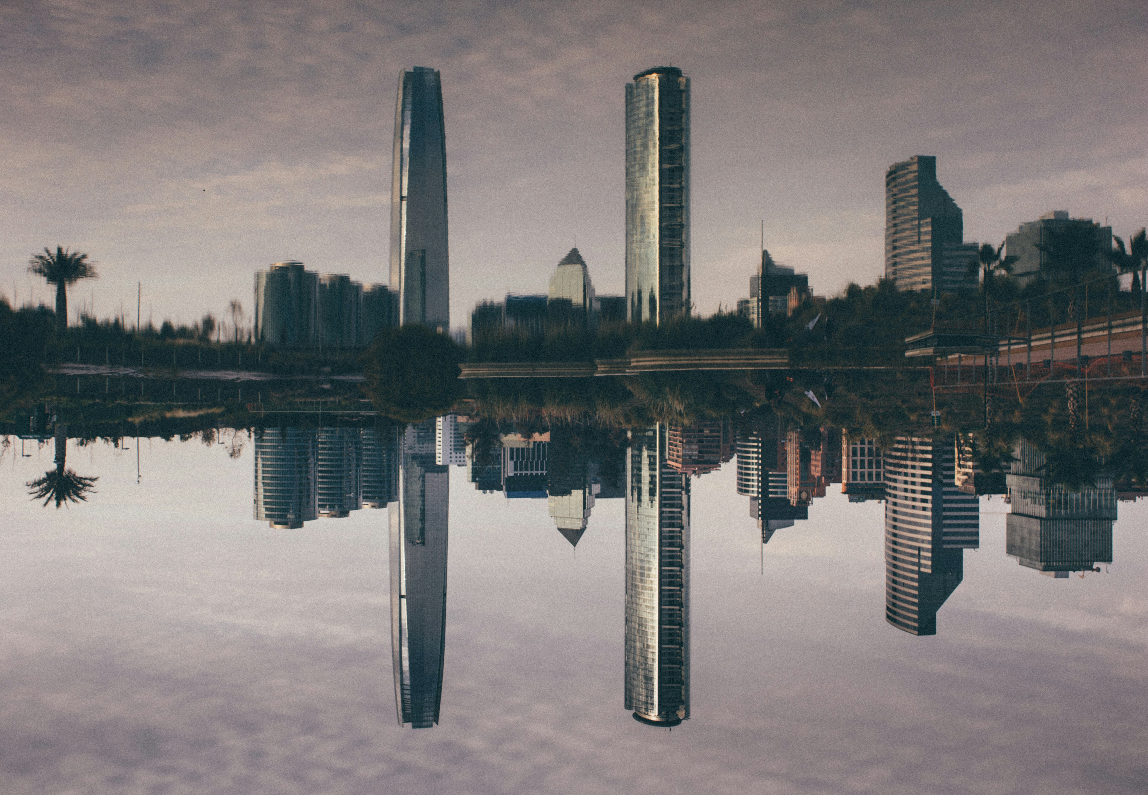 City skyline mirrored in a tranquil body of water, showcasing modern architecture against a soft, cloudy sky.