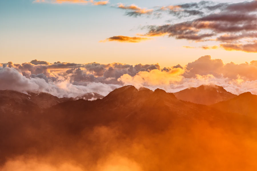 Dark mountain ridges rise above glowing clouds at sunrise, with warm orange light filling the sky and mist in the foreground.