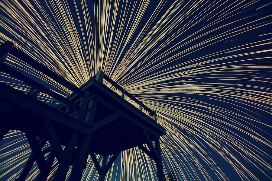 Long-exposure night scene capturing star trails above a silhouetted elevated platform, highlighting dynamic light streaks and architectural structure.