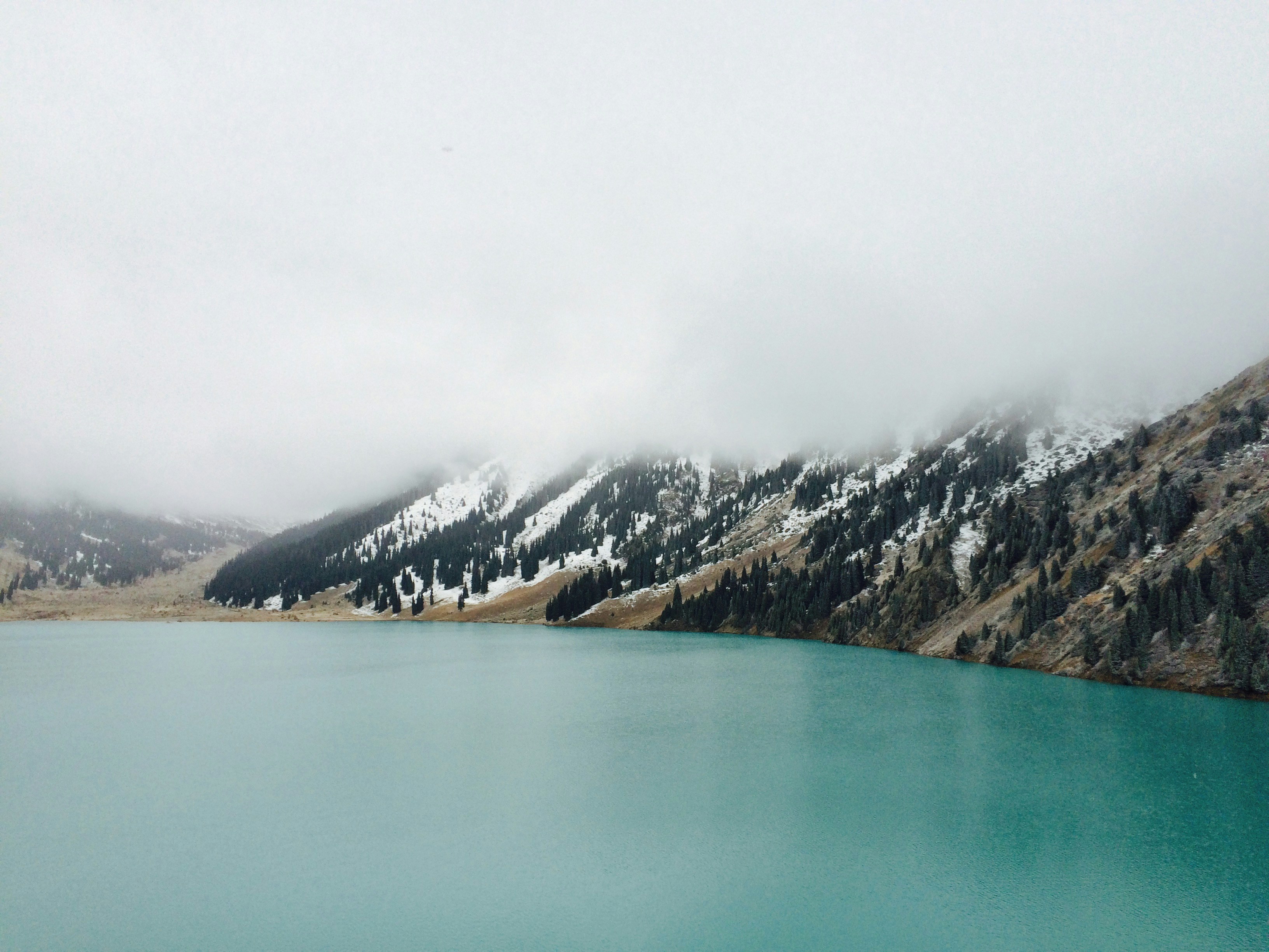 Turquoise mountain lake enveloped by dense mist, with snow-dappled peaks partially obscured.