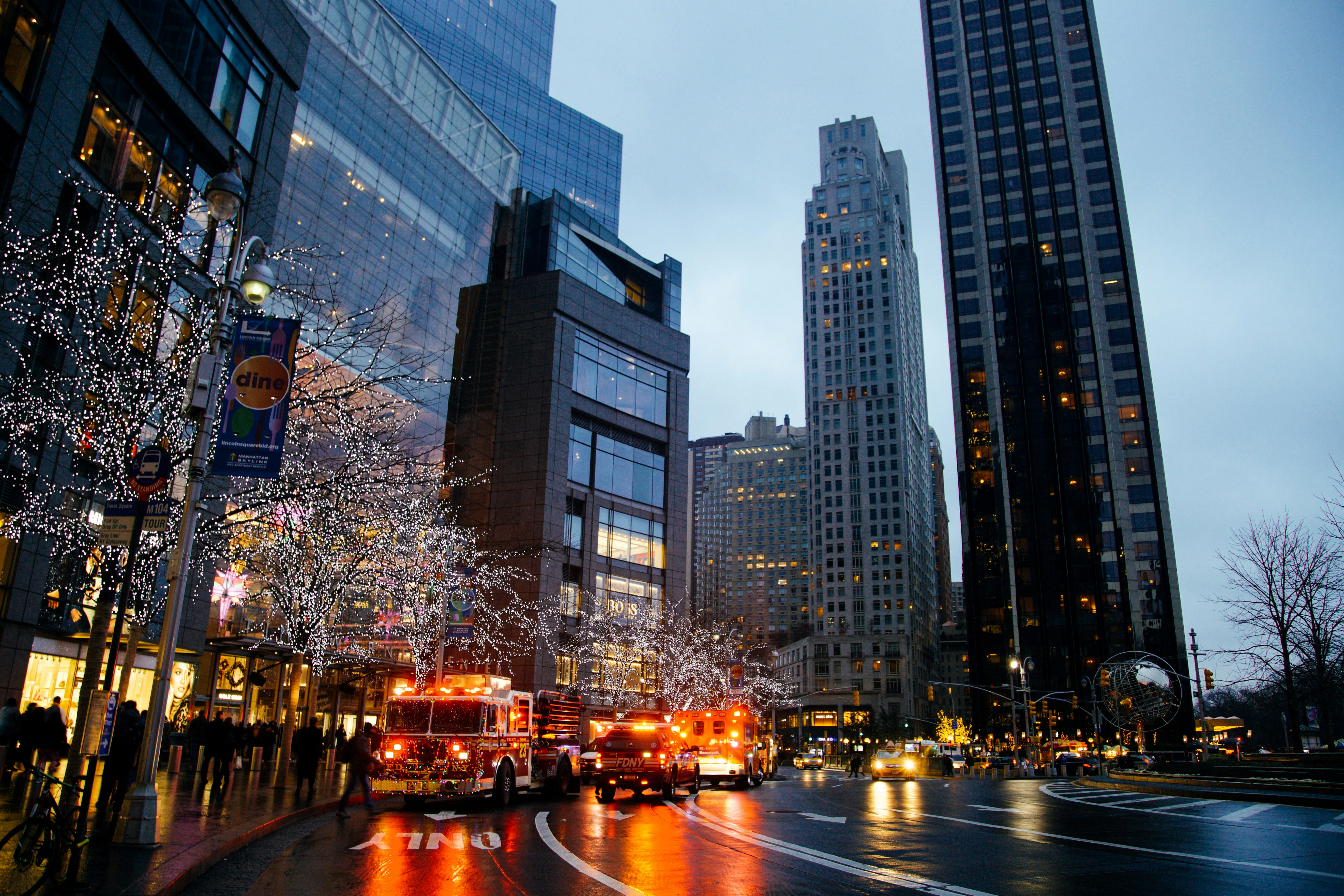 Fire trucks parked on a city street with trees adorned in white lights against a backdrop of tall buildings.