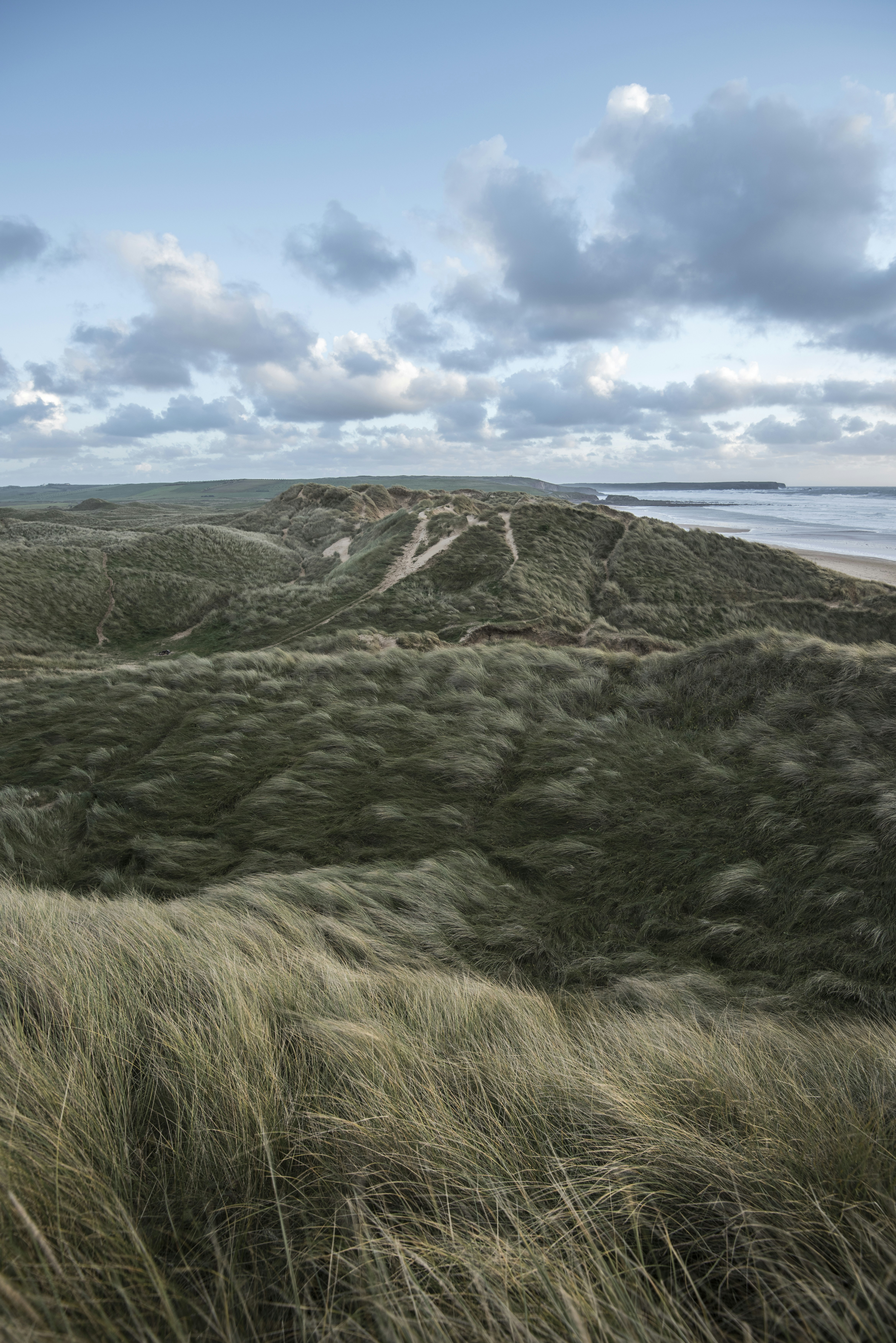 Rolling sand dunes covered in lush grass under a cloudy sky, leading to a serene beach in the distance.