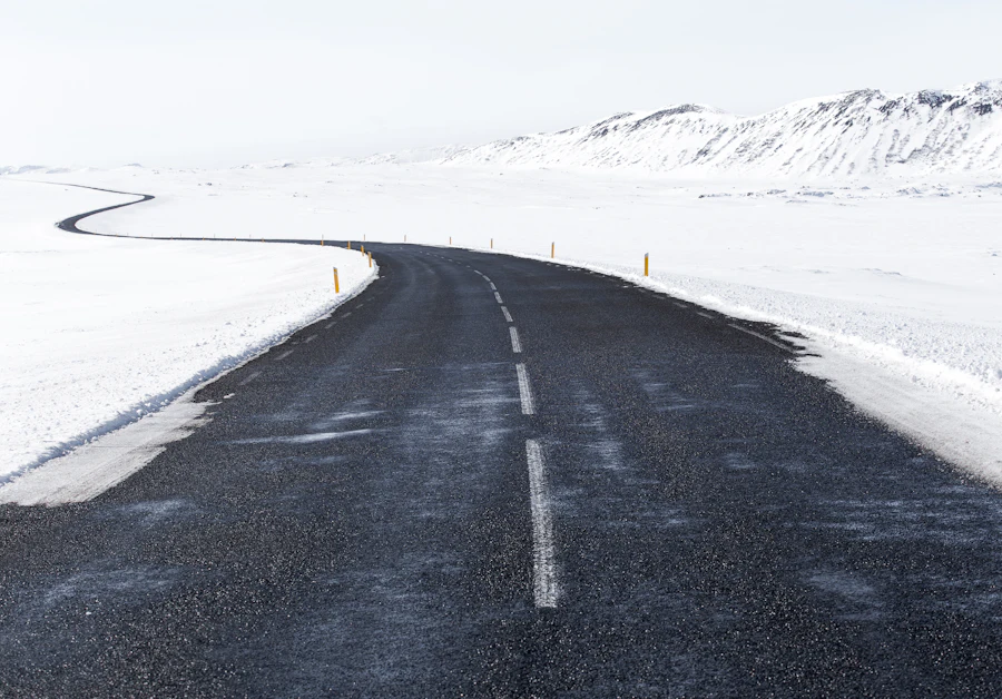 A dark asphalt road winds through a wide, snow-covered plain toward distant white hills. Yellow roadside markers punctuate the stark winter scene.