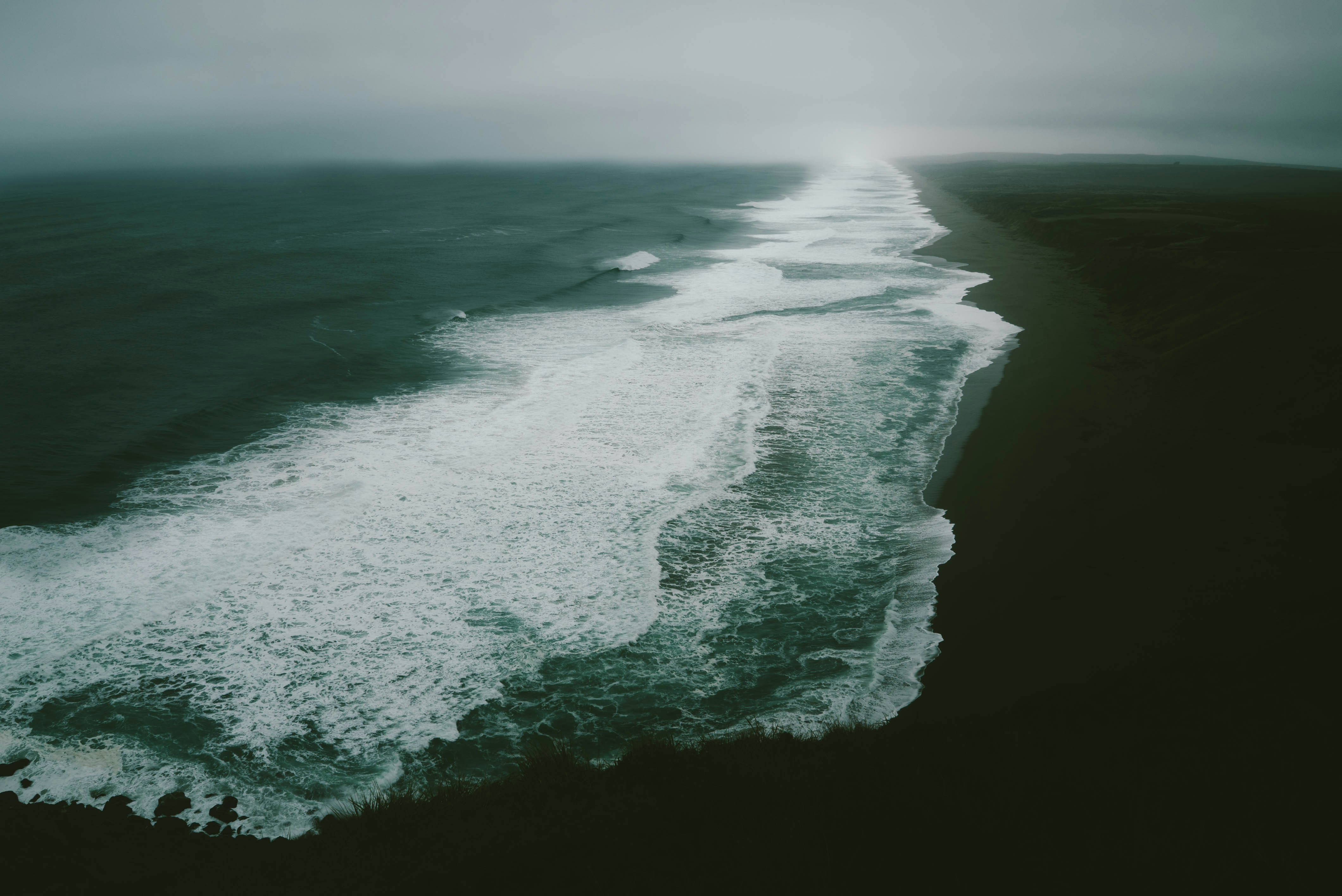 Waves crash along a dark shoreline under a moody sky, creating a dramatic contrast between sea and land. The horizon fades into the misty distance.