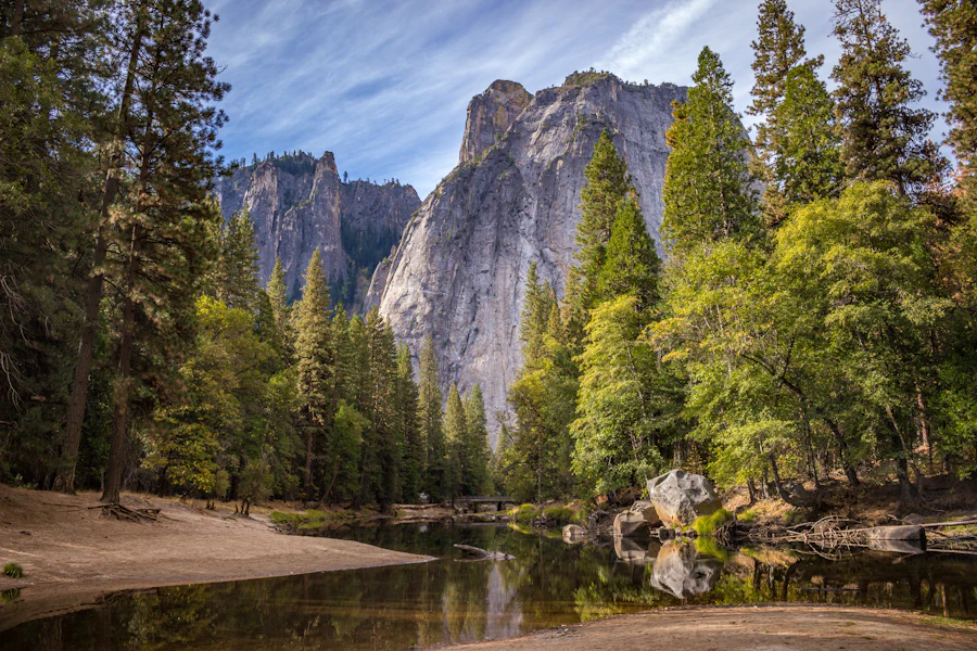 Towering granite cliff rises above a calm river framed by pine trees and dense forest. Soft clouds streak the blue sky, with the rock face reflected in the water.